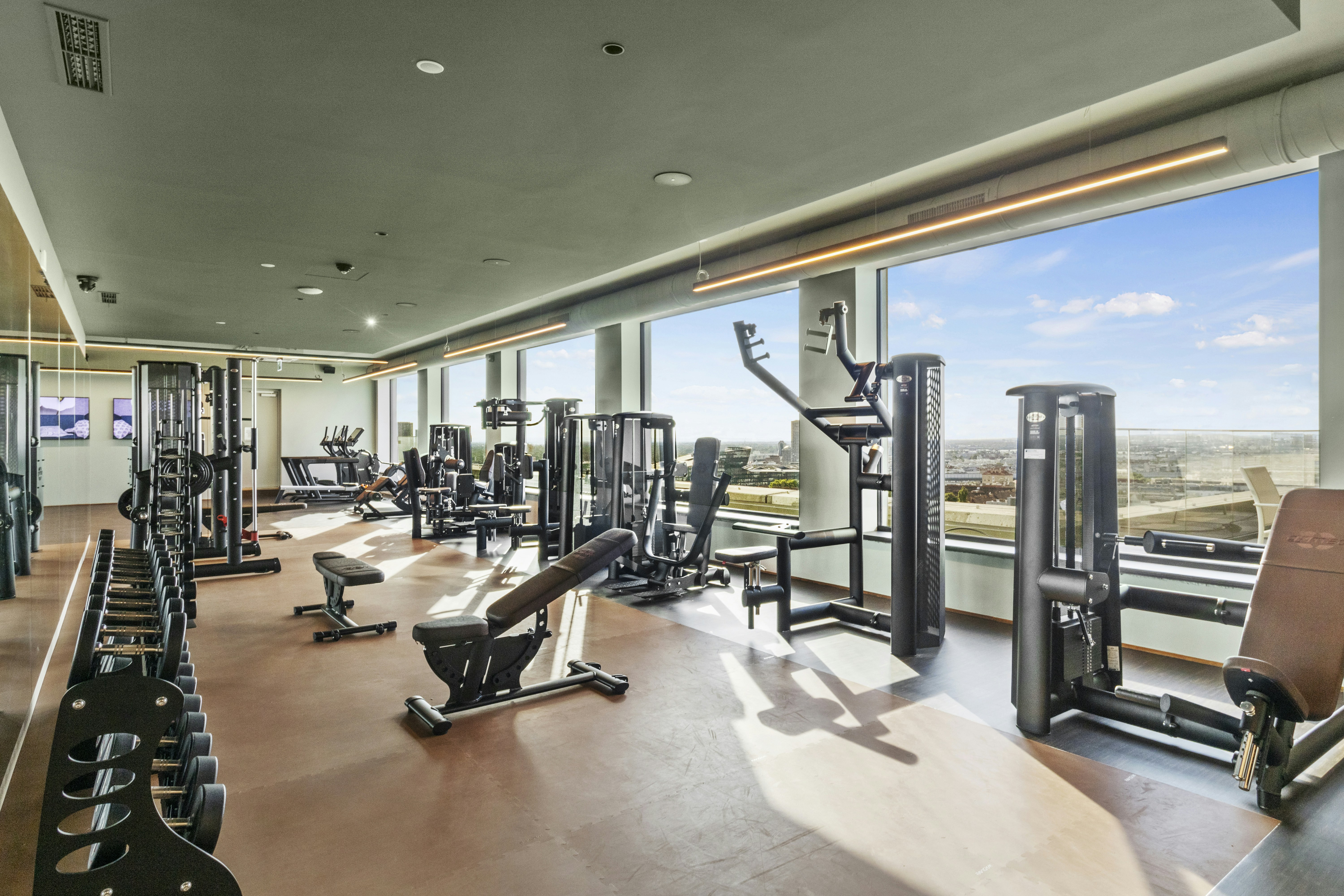 Bright hotel gym with large windows, cardio and weight machines, and a view of the city skyline; concrete floor and recessed ceiling lights. The scene captures a spacious fitness space designed for functional training.