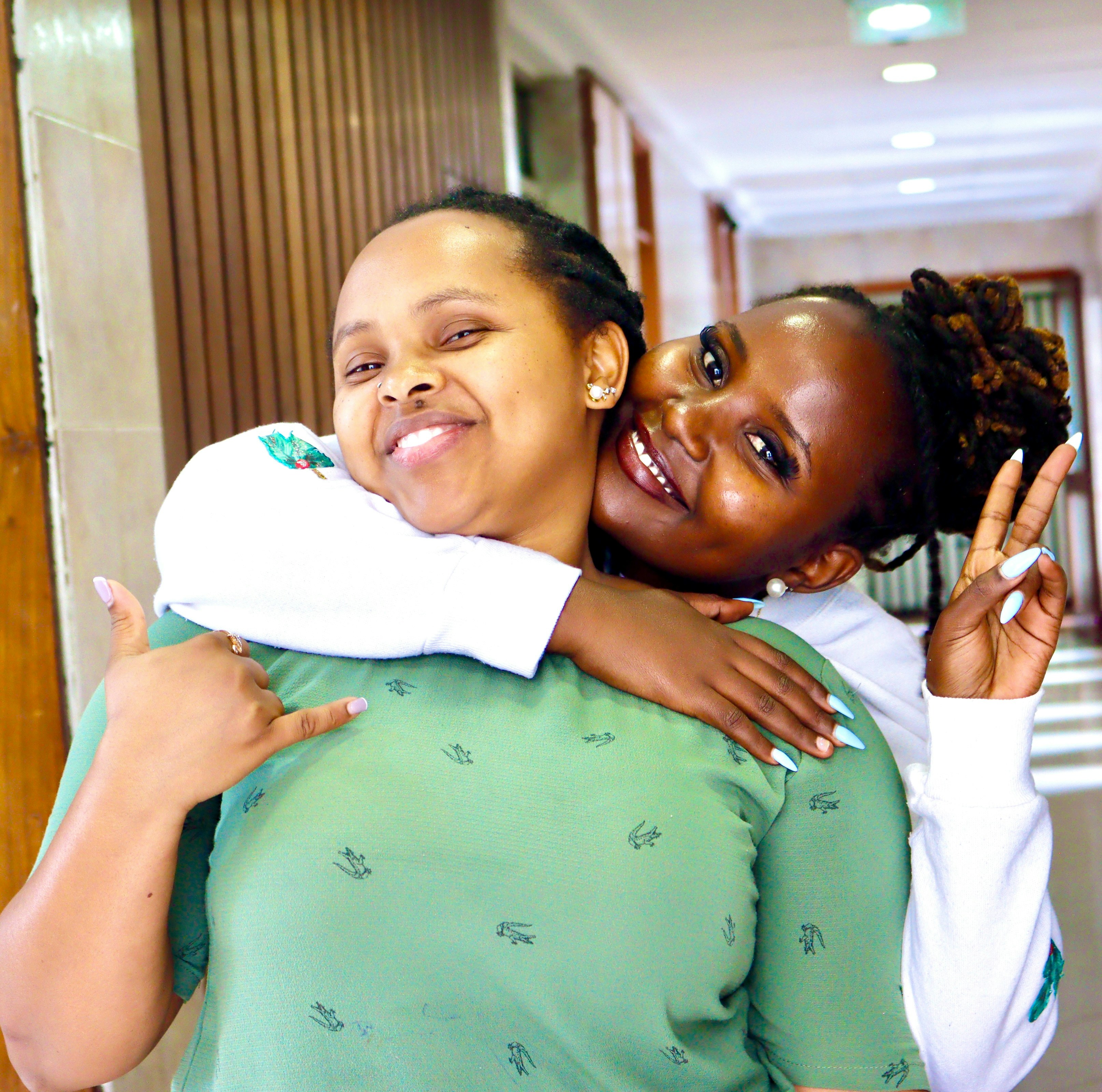 Two friends share a joyful hug in a bright hallway; the moment centers on their faces and smiles, with one making a playful gesture. A candid, friendly portrait captured in natural indoor light.
