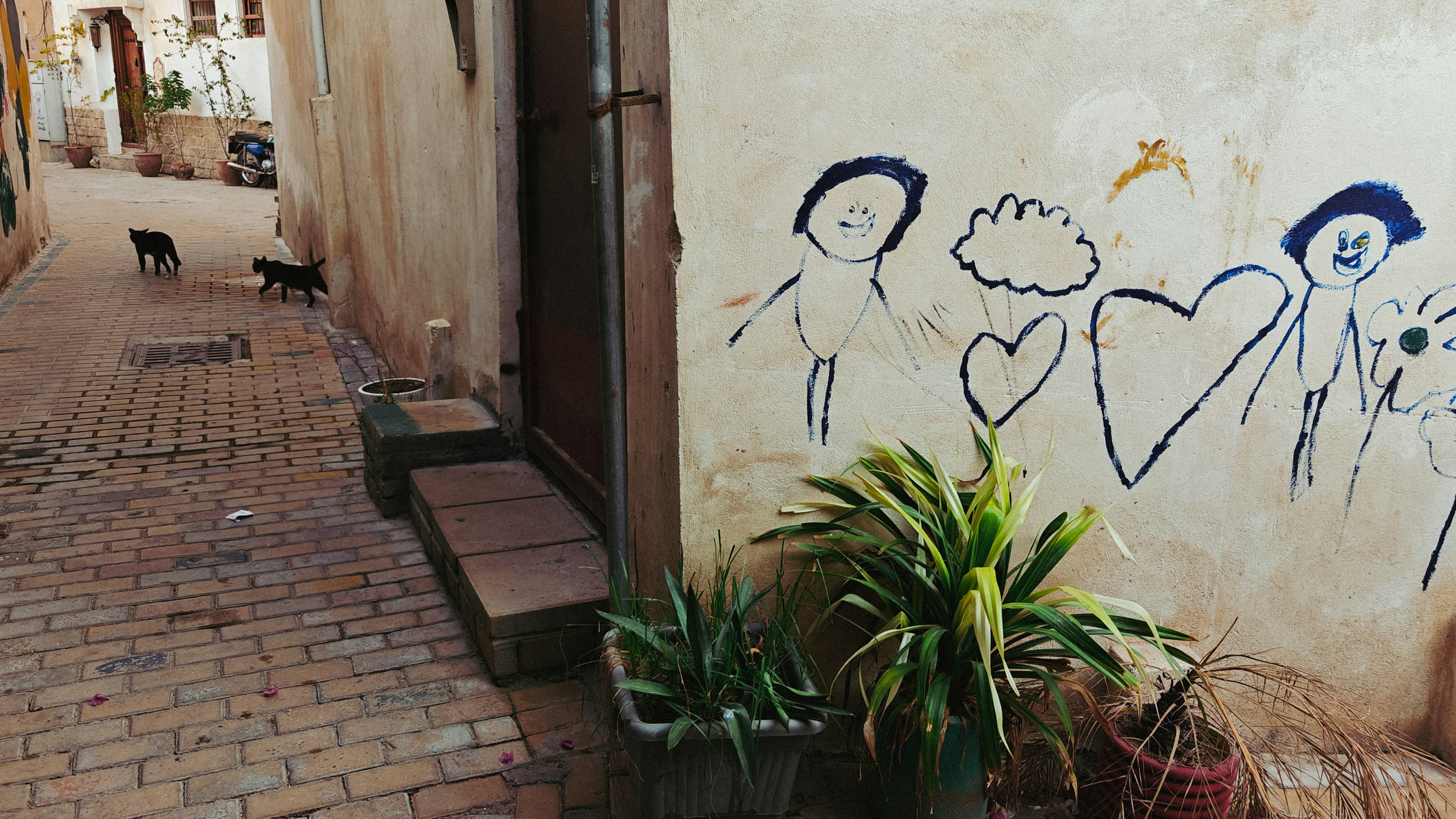 Narrow brick alley with distant cats and a mural featuring childlike figures and hearts on a sunlit wall.