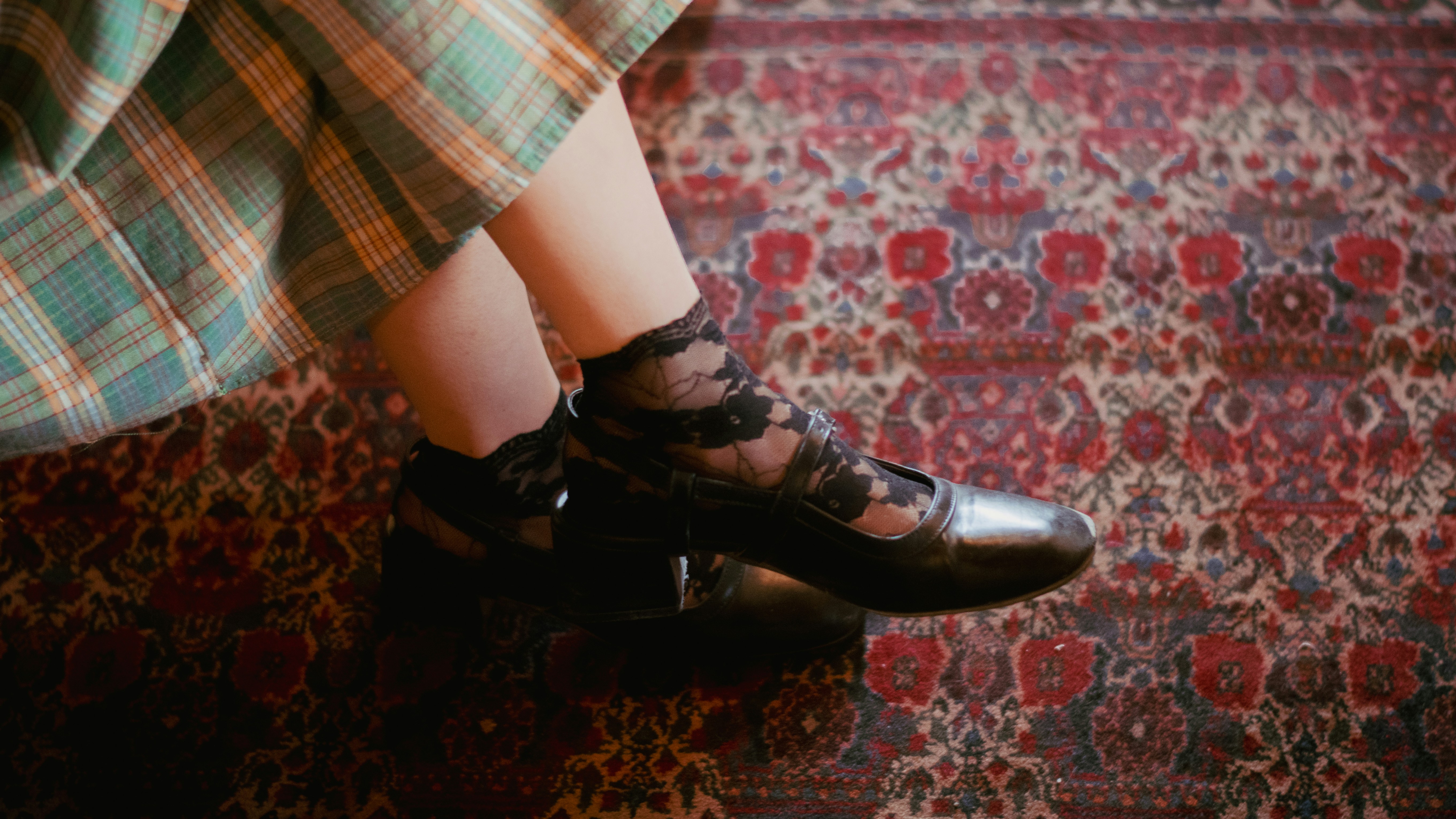 Pair of black lace socks and Mary Jane shoes sit on a richly patterned red rug, with a plaid skirt draped nearby; composition emphasizes vintage footwear and texture of the carpet.