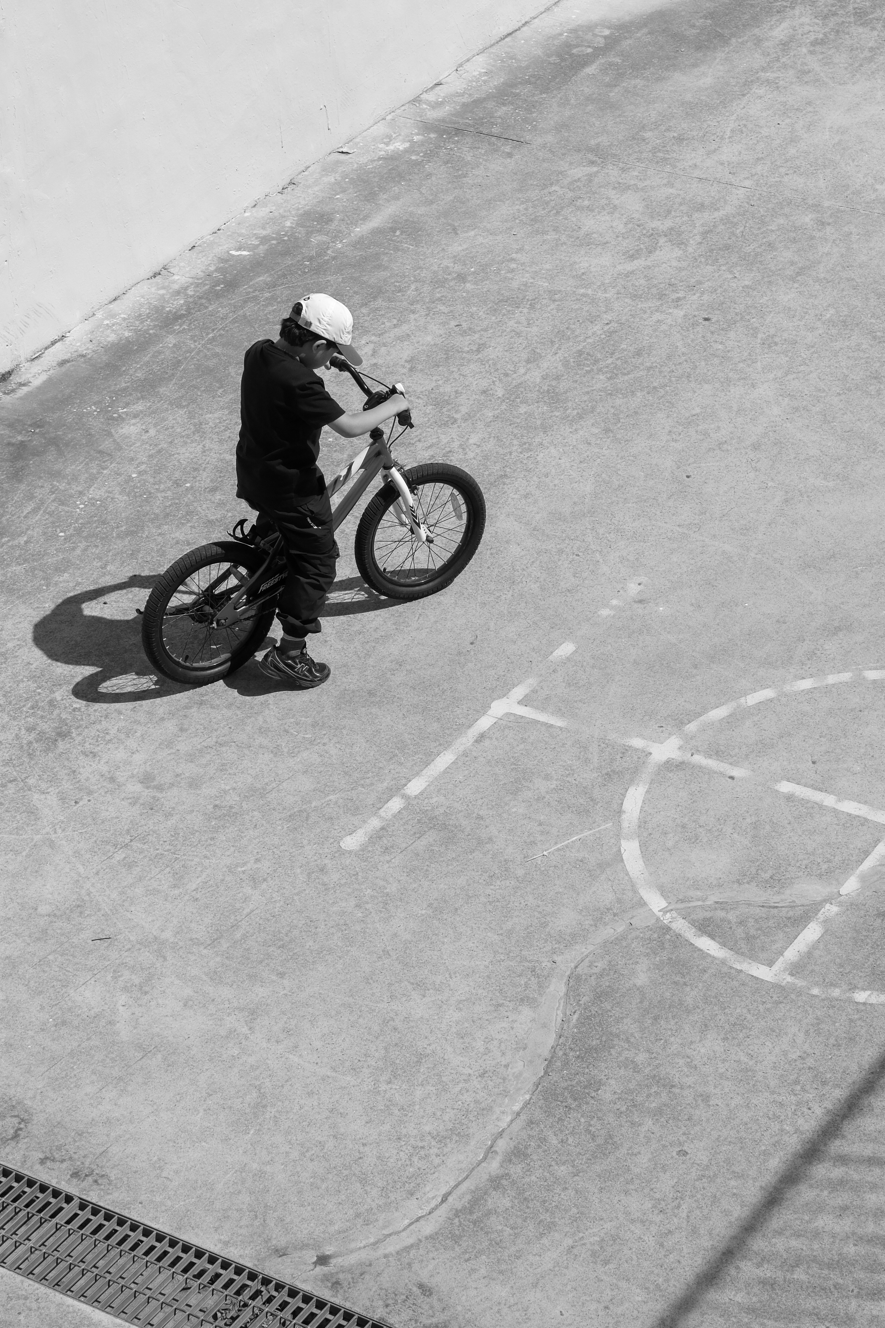 A lone BMX rider sits paused on a sunlit concrete court, shadows stretching across the textured surface with chalk markings nearby.
