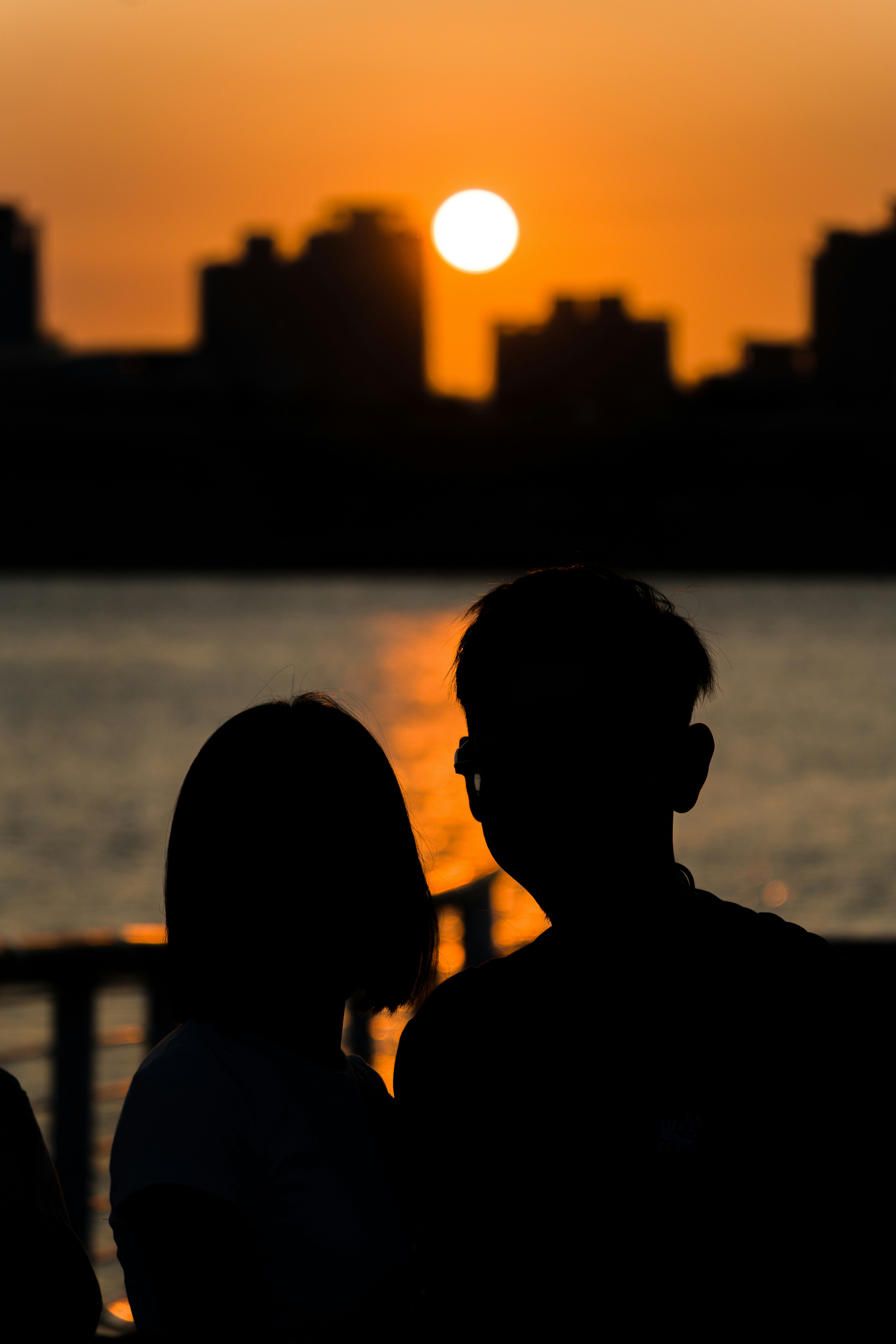 Silhouetted couple gazing toward a setting sun over a waterfront, with city skyline silhouettes in the background. A minimal scene focusing on mood and ratio of light at golden hour.