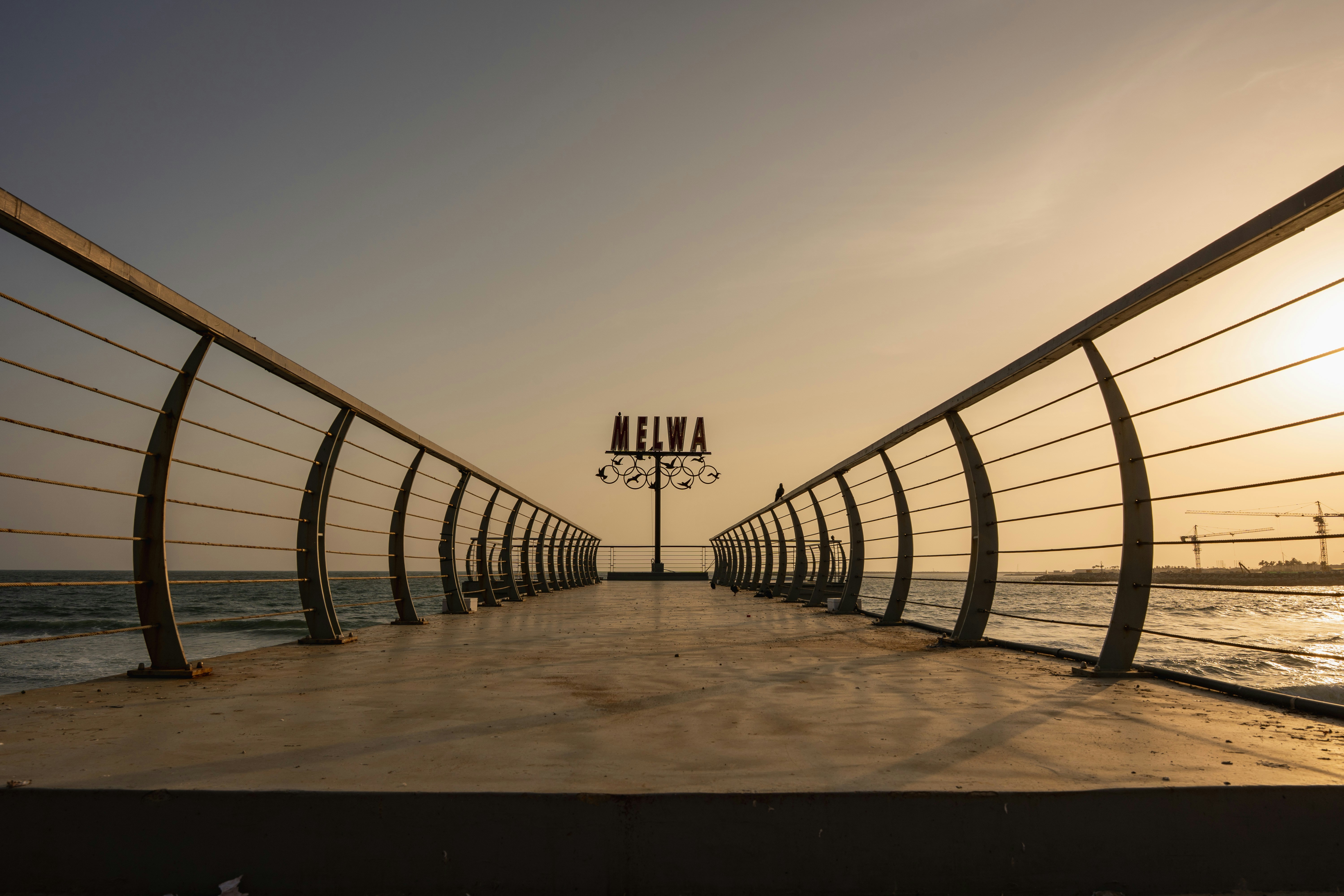 A concrete pier with metal railings leading to a sign reading "MELWA" at sunset