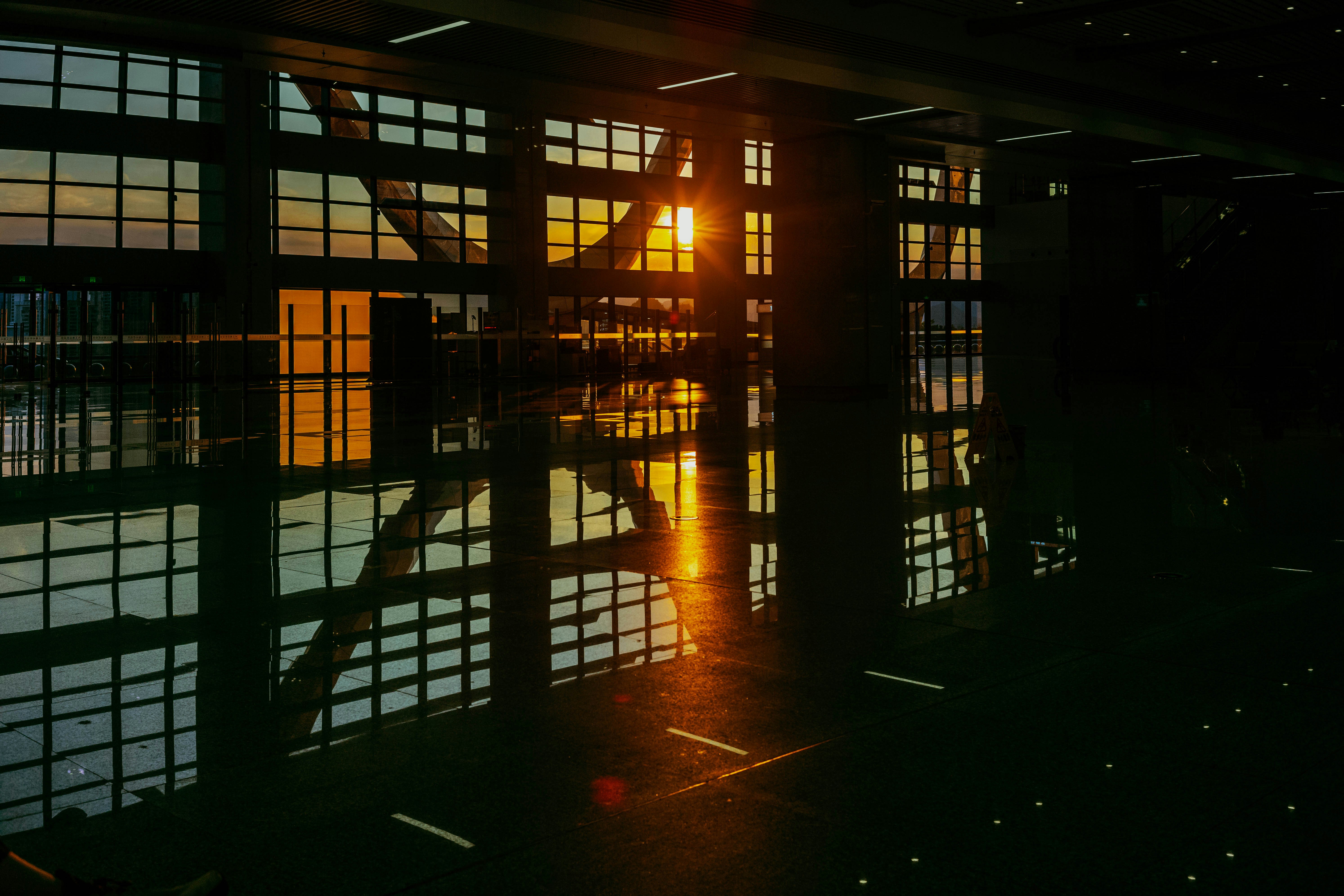 Sunlight streams through industrial windows, creating a warm glow and reflective patterns on the dark interior floor. A silhouetted structure and water-like reflections emphasize the contrast between light and shadow, with a moody, architectural mood.
