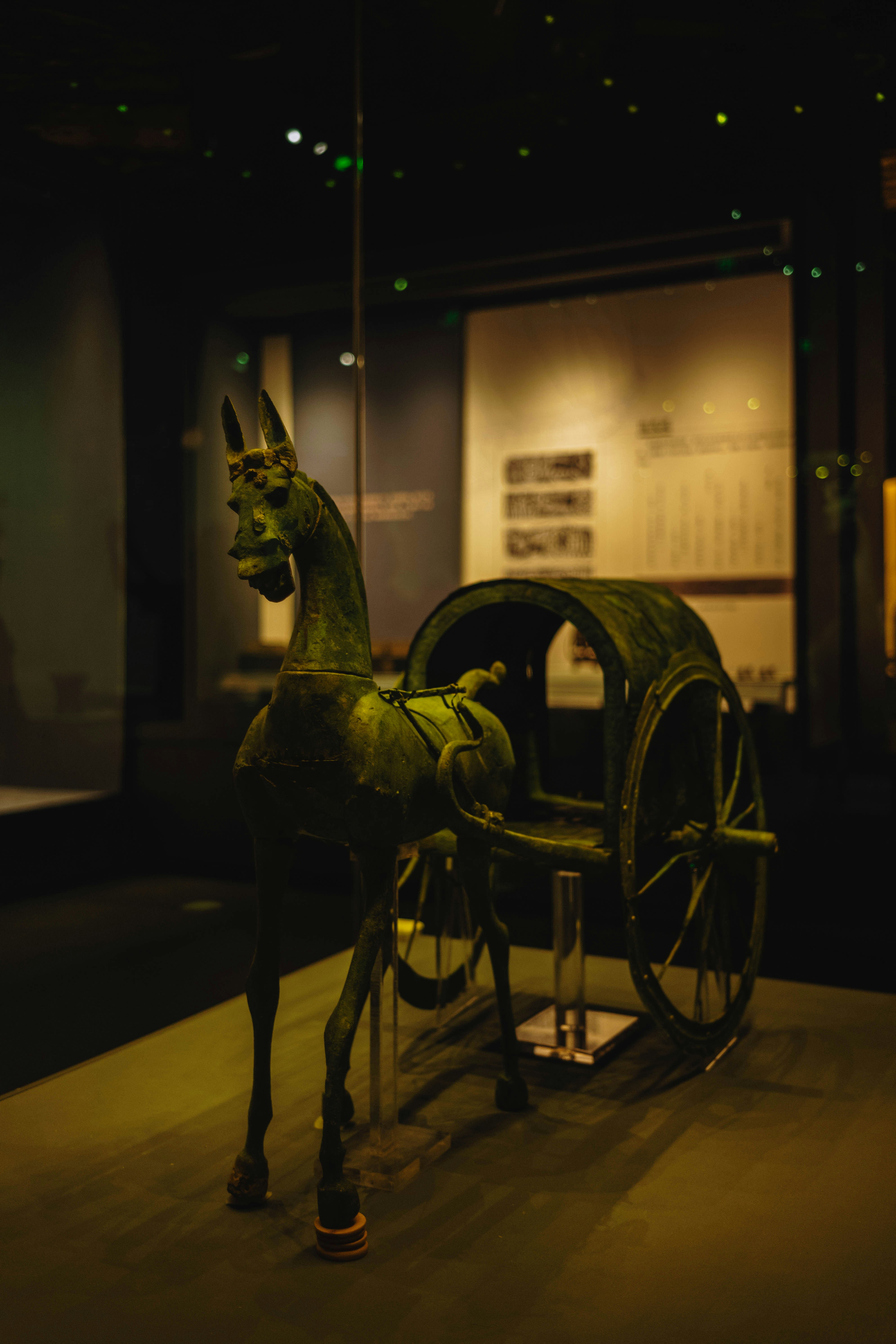 Bronze horse and cart on display in a dim museum case, illuminated by warm ambient lighting.