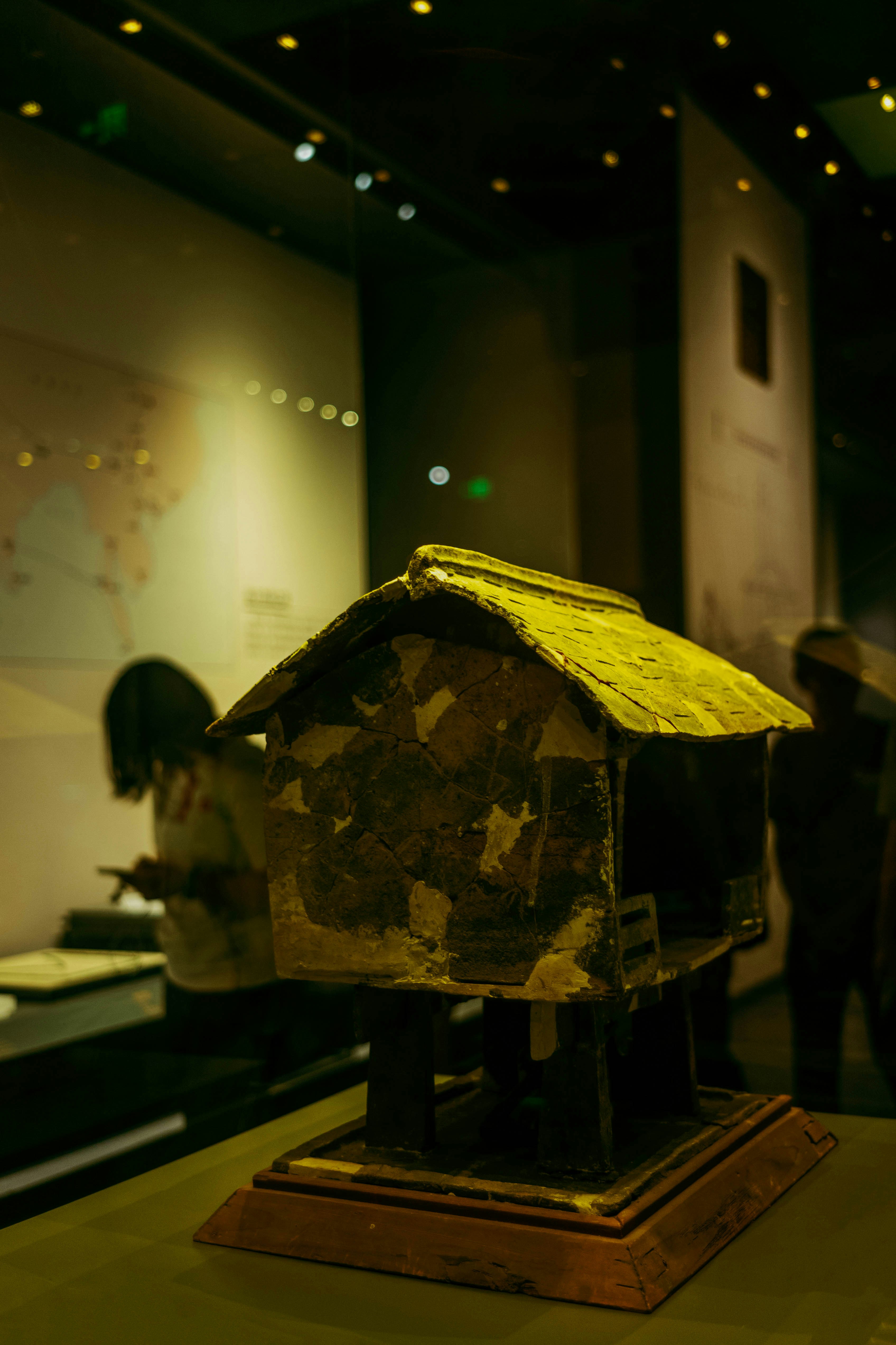 A small weathered model house on a wooden stand sits under museum lighting, with blurred visitors in the background.
