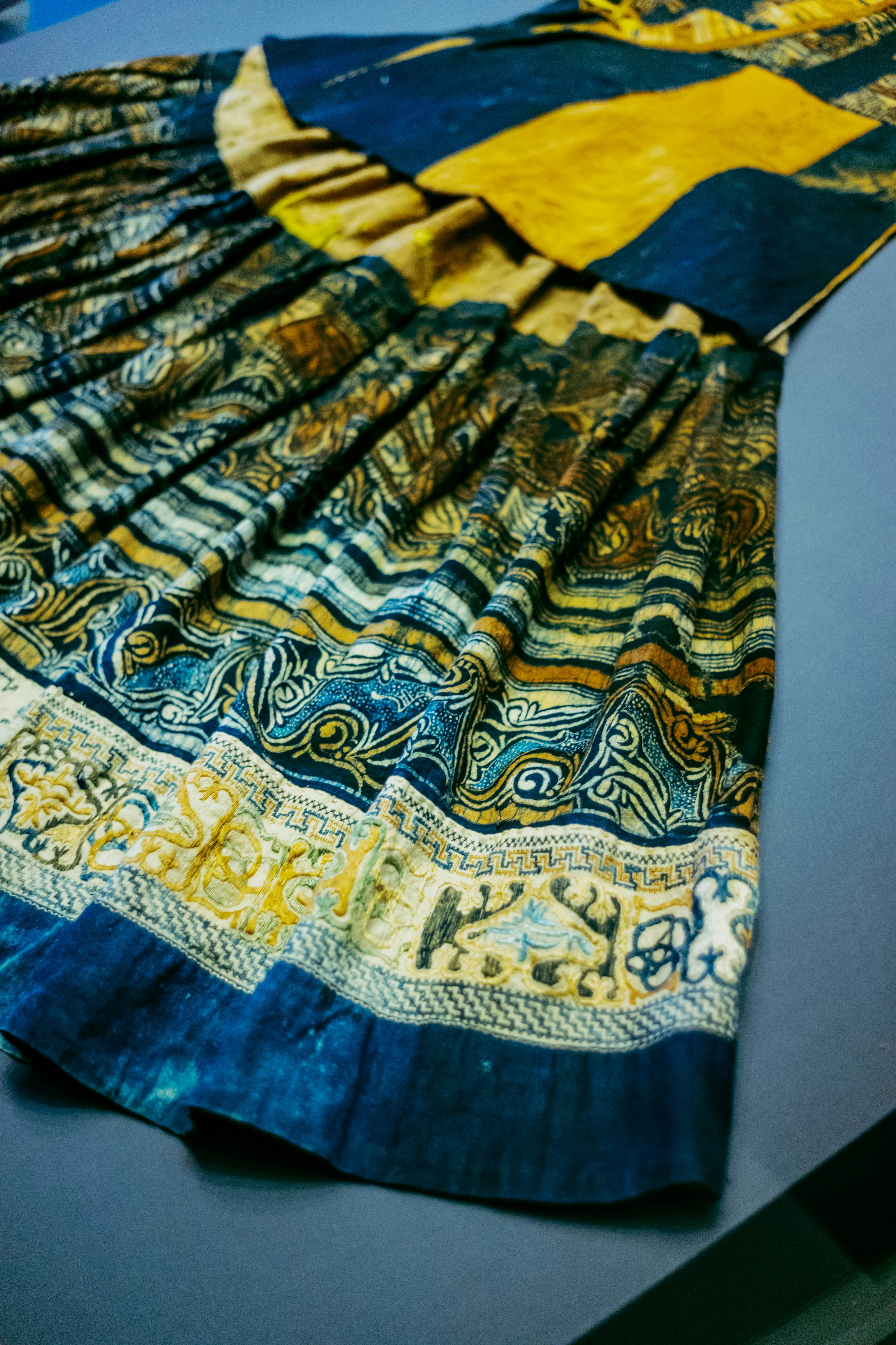 Textured fabric with intricate decorative patterns in yellow, blue, and gold tones draped diagonally; a close-up study of weave and color.