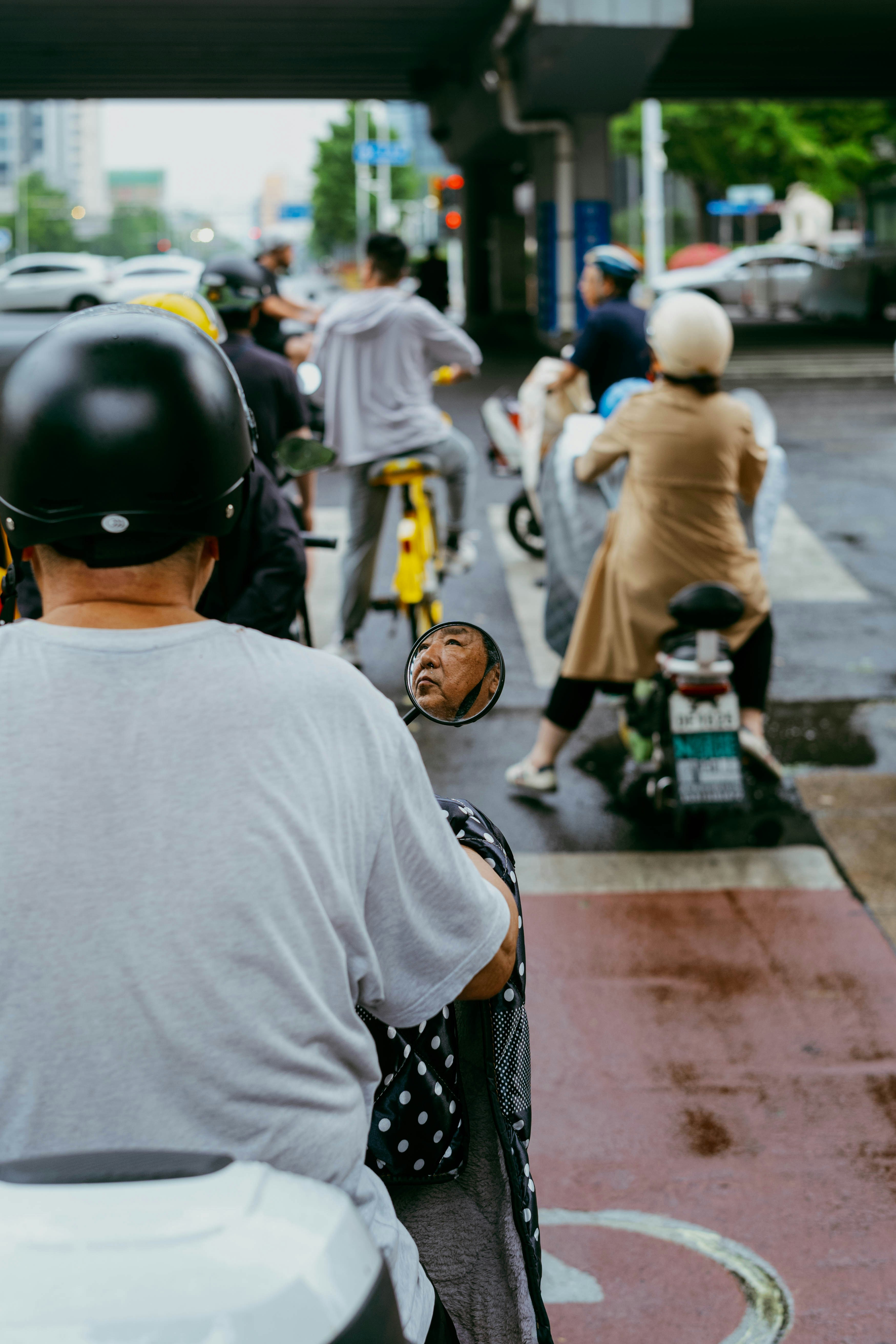 Urban traffic scene with a rear rider looking into a small round rear-view mirror, amid scooters and pedestrians at a crosswalk.