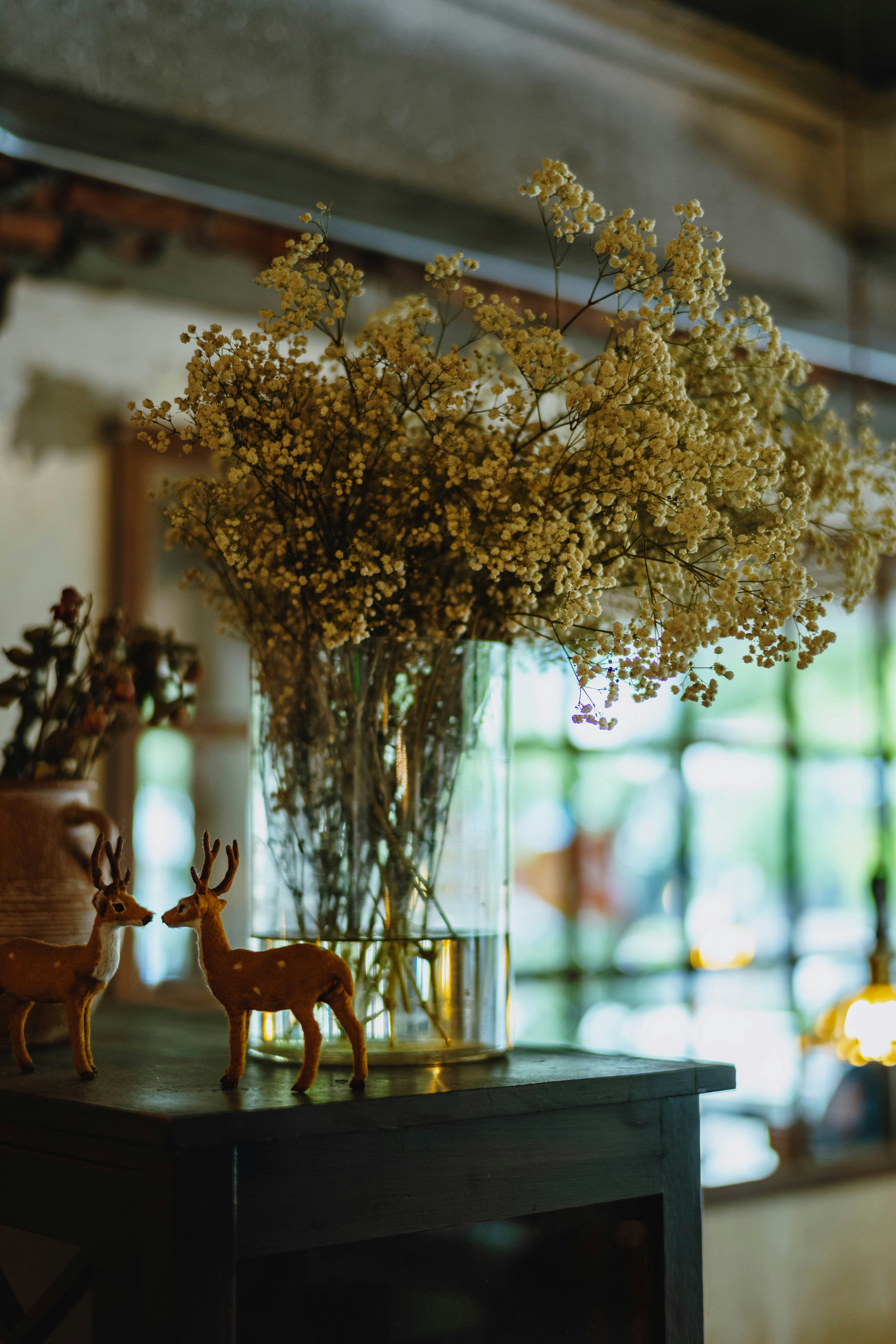 A glass vase with dried yellow blossoms sits on a dark wooden shelf beside two small deer figurines, with a softly blurred background suggesting a cozy interior scene.