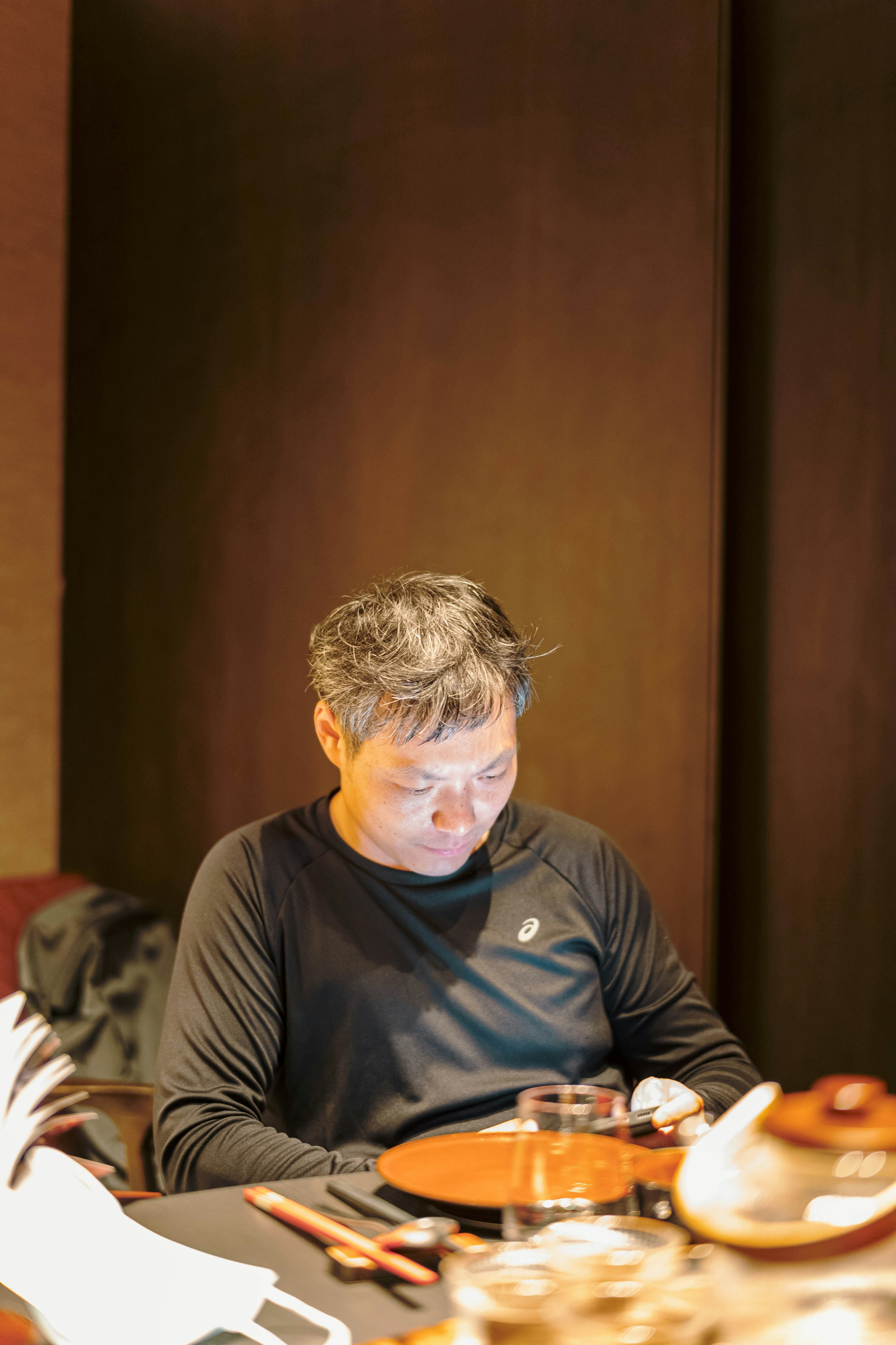 Portrait of a man seated at a dining table, looking down with warm ambient lighting and a shallow depth of field. A candid, intimate moment captured in a dinner setting; the shot emphasizes the subject against a softly blurred background.