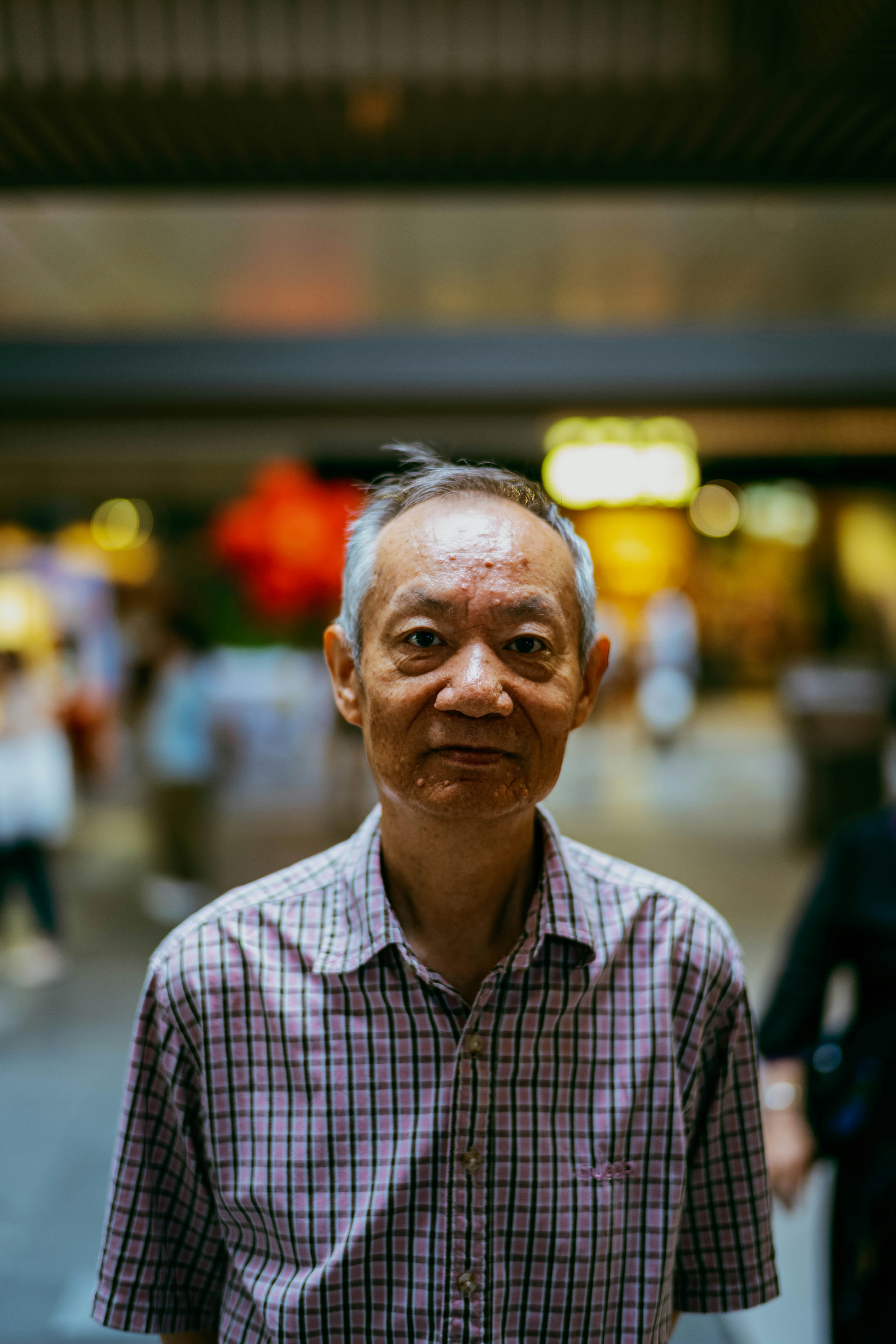 Elder man stands facing the camera in a busy urban interior, softly lit by background neon and bokeh-filled scenes.