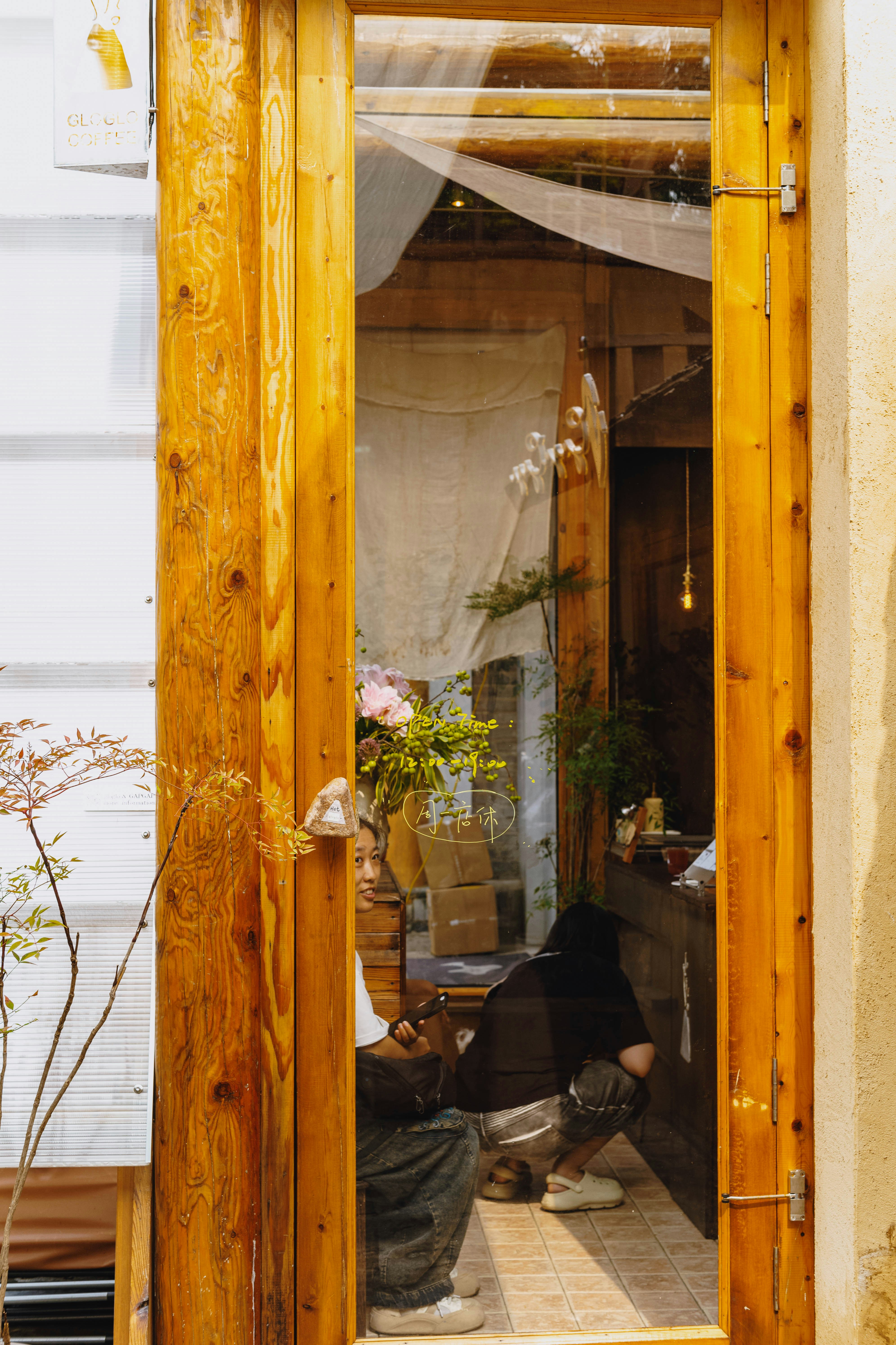 Softly lit interior seen through a tall wooden-framed glass door; two people seated inside, one facing the camera, with floral arrangements and warm tones inside a cozy café.