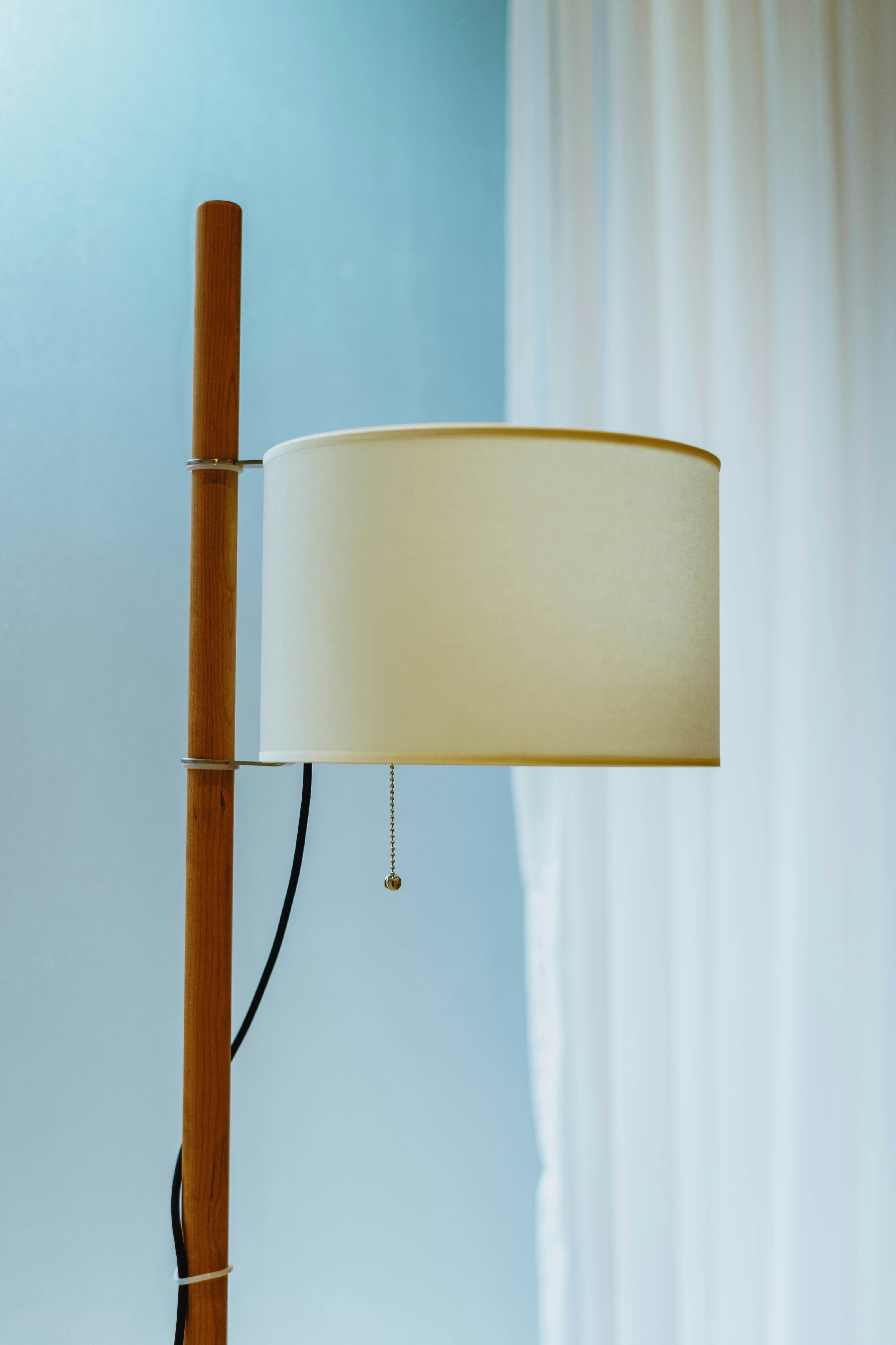 Sleek mid-century lamp with a fabric shade mounted on a vertical wooden pole against a pale blue wall and sheer white curtains.