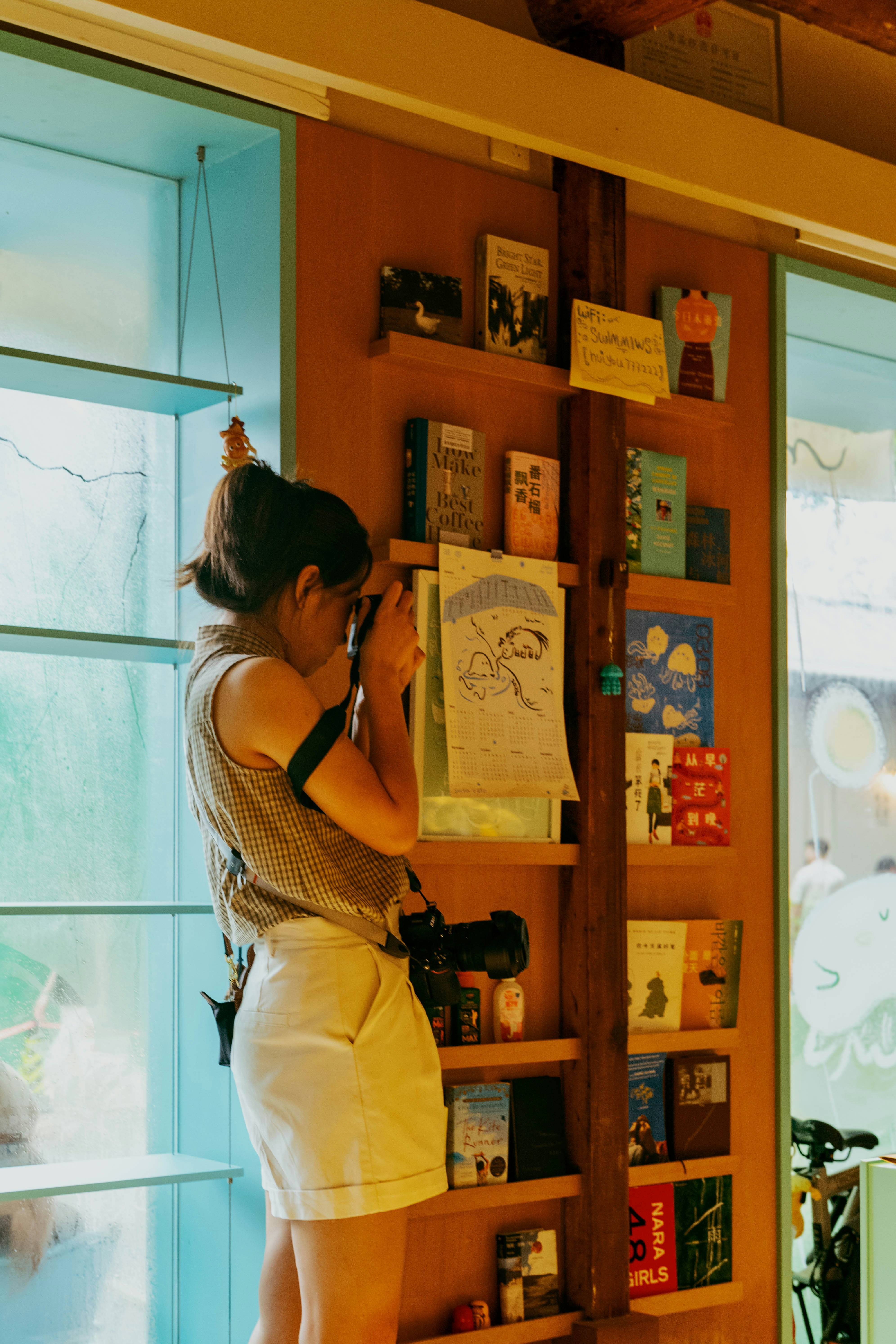 A woman with a camera stands inside a cozy bookstore, adjusting her lens while surrounded by shelves of colorful books. The scene emphasizes a candid moment of creative work in a warm, inviting space.