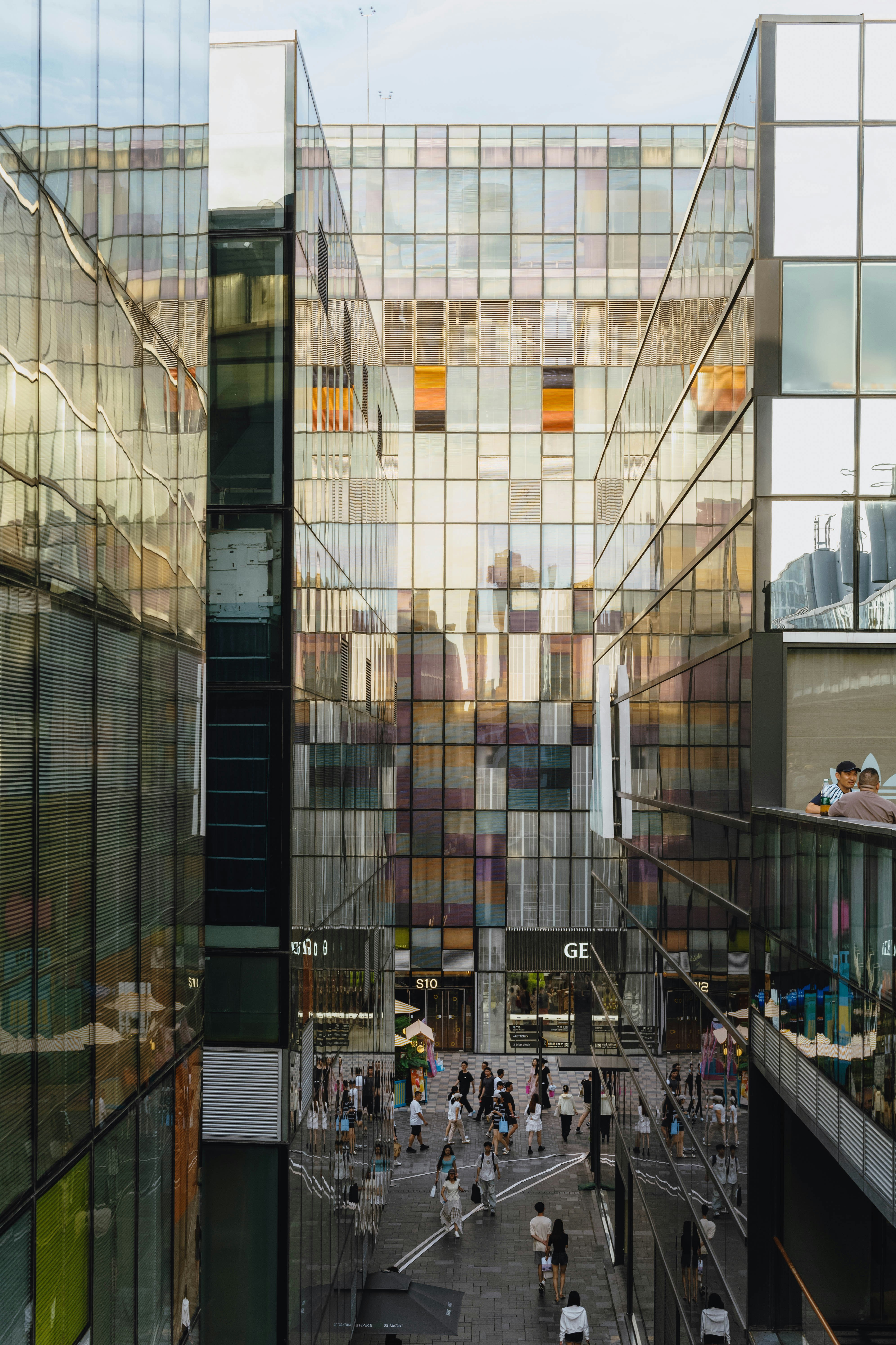 A bustling urban plaza framed by mirrored glass buildings, with pedestrians moving through a sunlit courtyard. The image captures the reflective architecture and everyday activity within a vertical, canyon-like atrium.