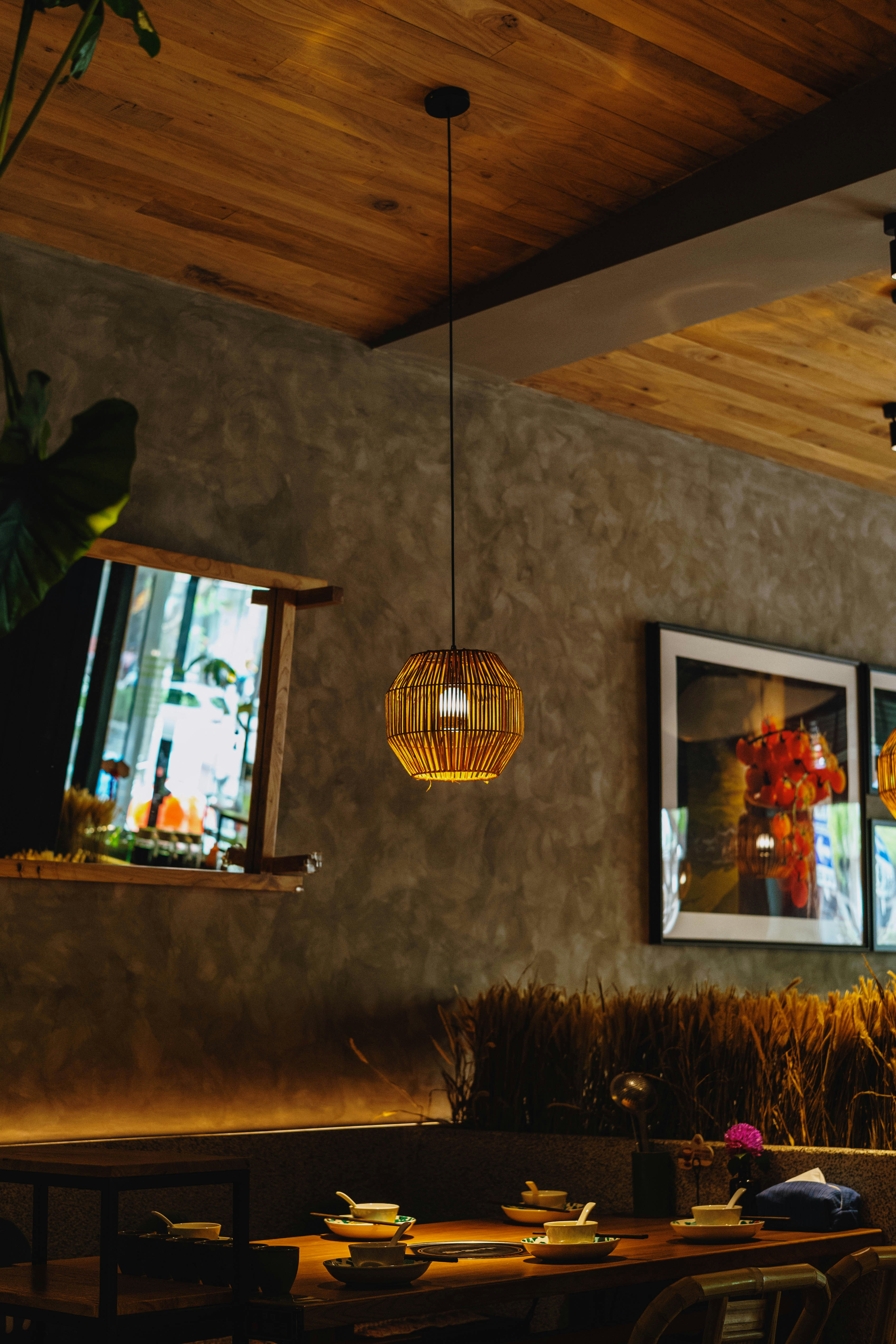 Warm, dimly lit restaurant interior with a hanging wicker lamp centered above a wooden table set for dining; framed art on the textured wall and a plant to the left add depth to the scene.
