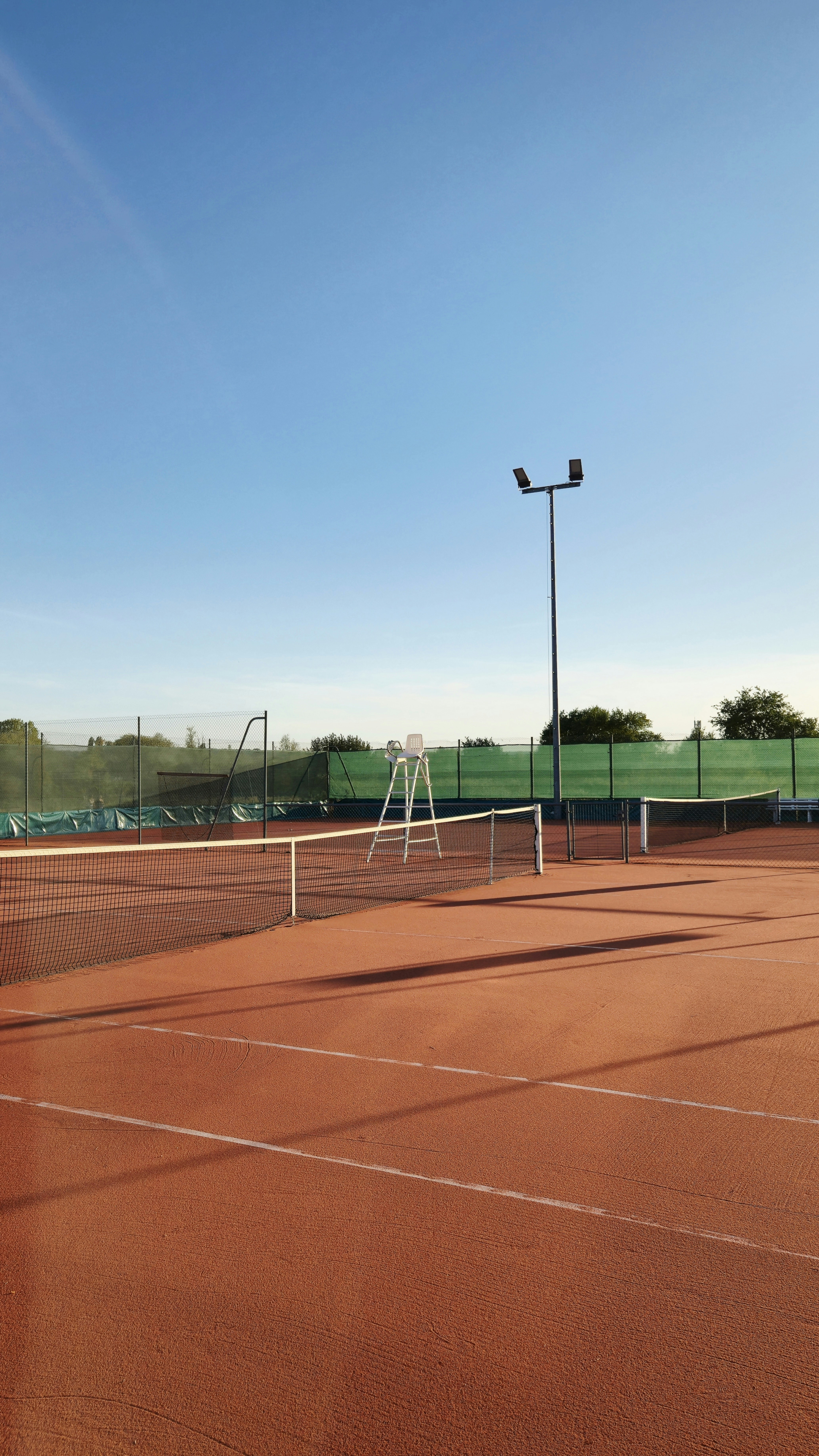 A clay tennis court with a white umpire chair under a clear blue sky