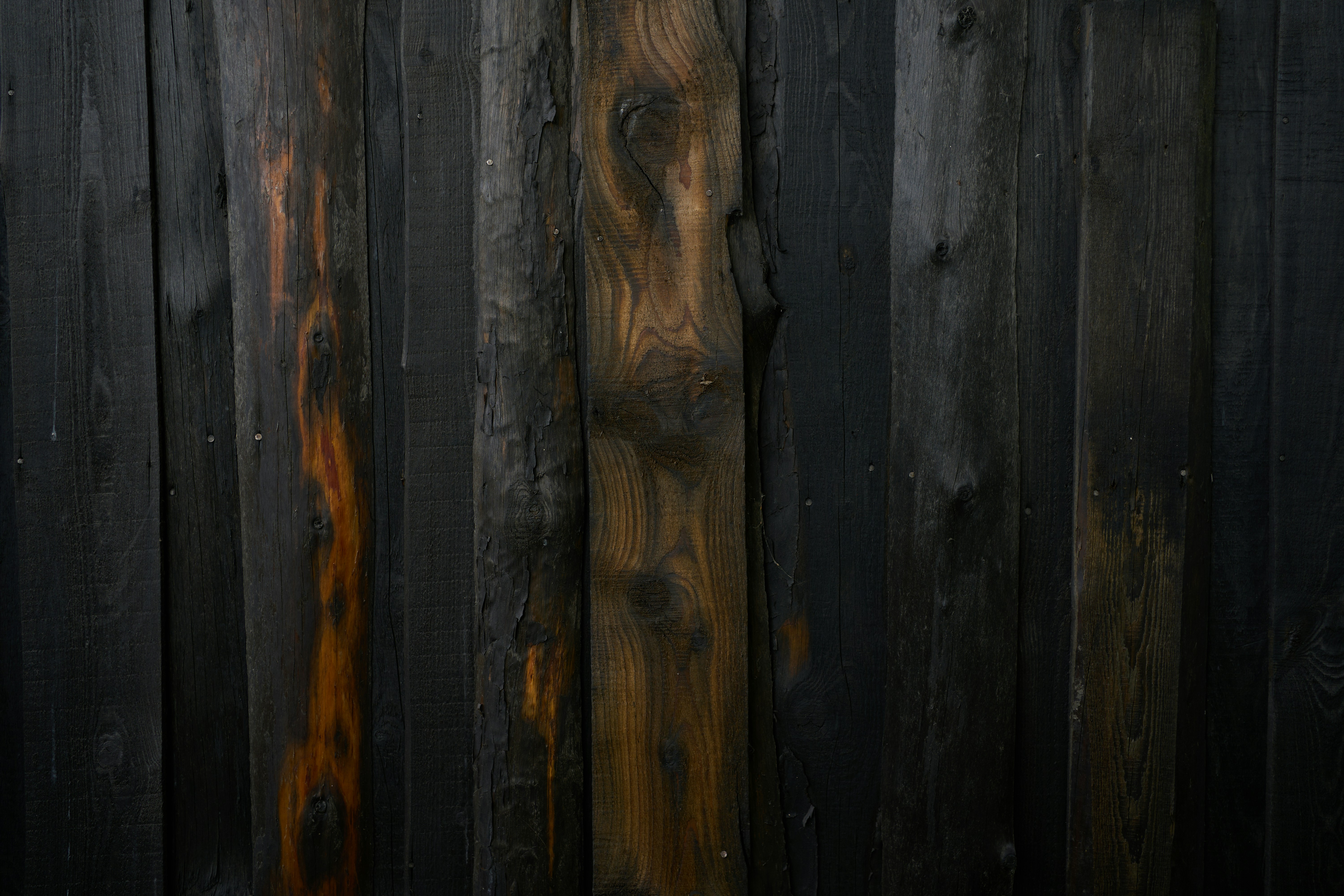 Close-up of vertically aligned, weathered wooden slats with dark, charred tones and subtle orange-brown highlights; a moody architectural detail with rich texture and grain.