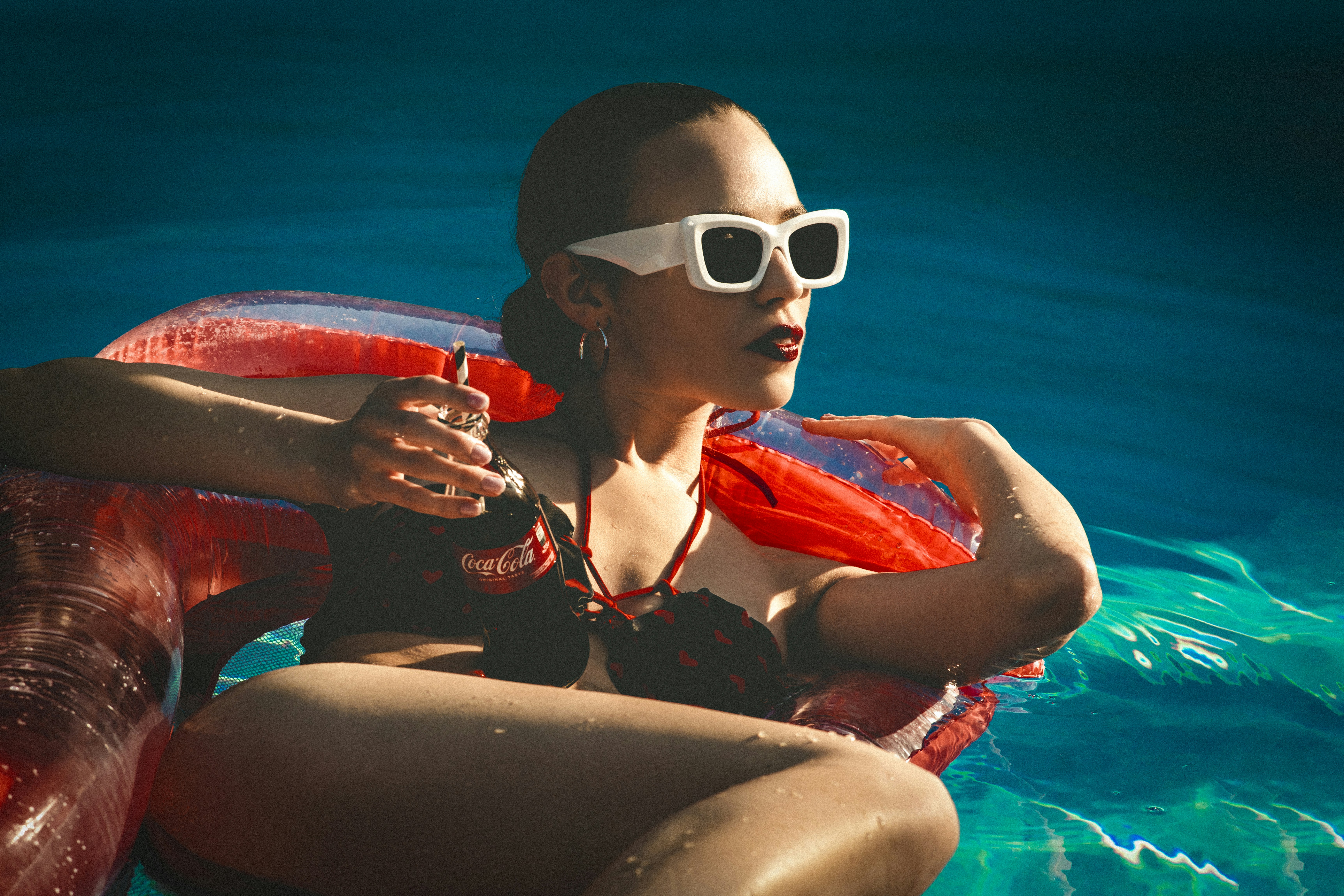 Woman lounging on a red inflatable float in a bright pool, wearing white sunglasses and a black bikini; Coca-Cola bottle in hand. This is a lifestyle, candid-like photograph capturing relaxed summer leisure.