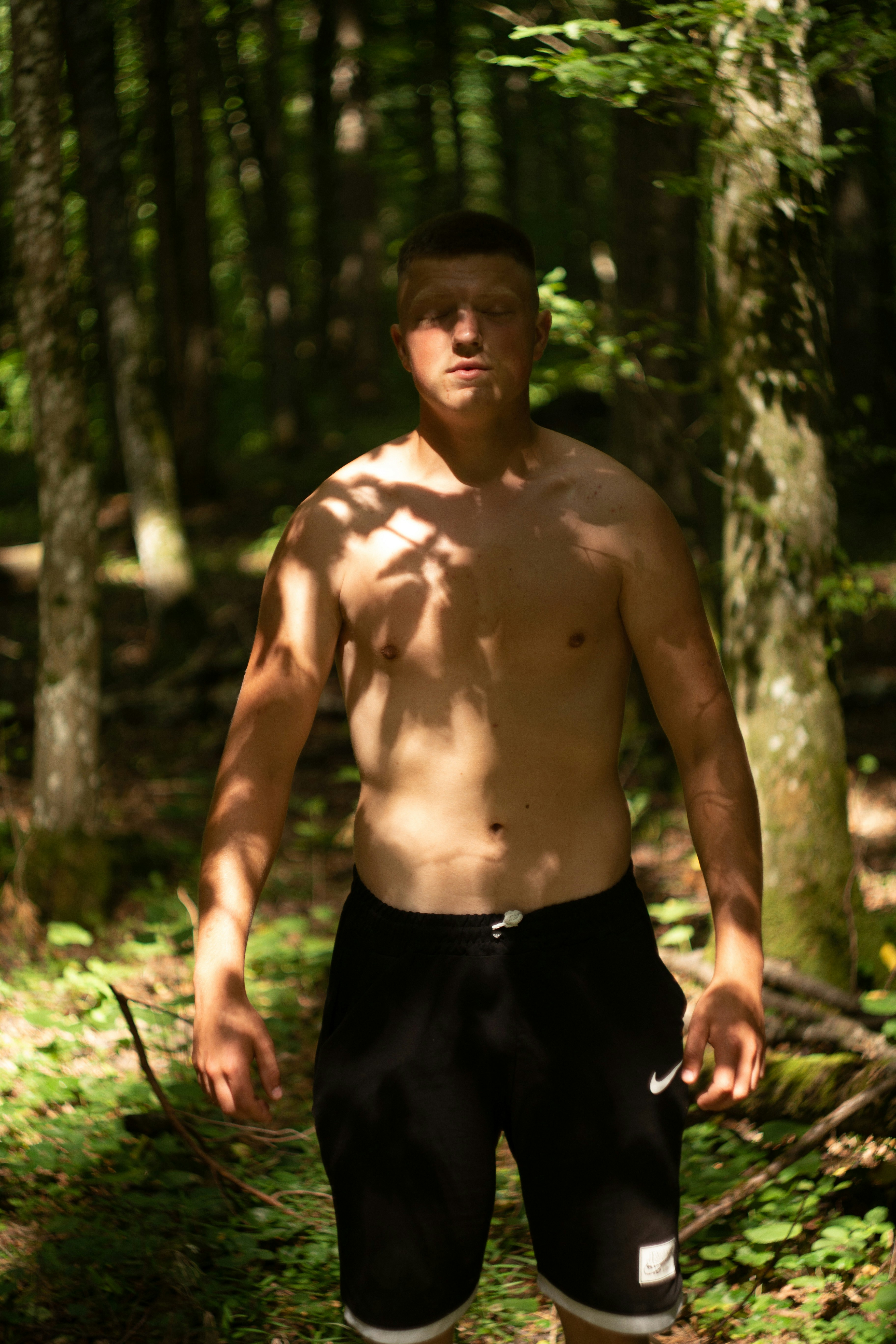 Shirtless man standing in a sun-dappled forest clearing, wearing black shorts; soft natural light creates mottled shadows across his torso while trees form a green backdrop.