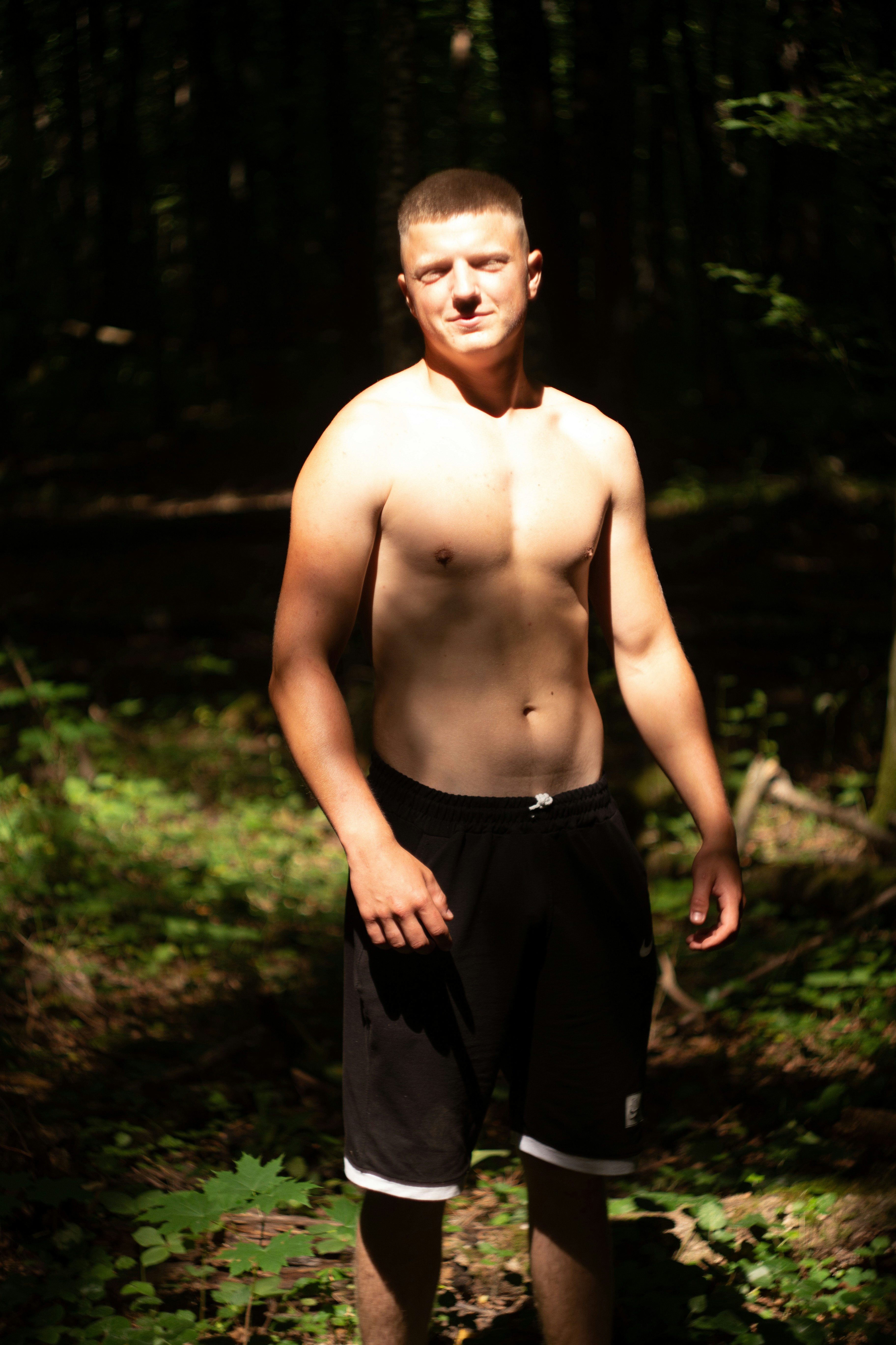 A shirtless man stands in a sun-dappled forest clearing, the torso and face highlighted while background falls to shadow; a natural outdoor portrait emphasizing physique and environment.