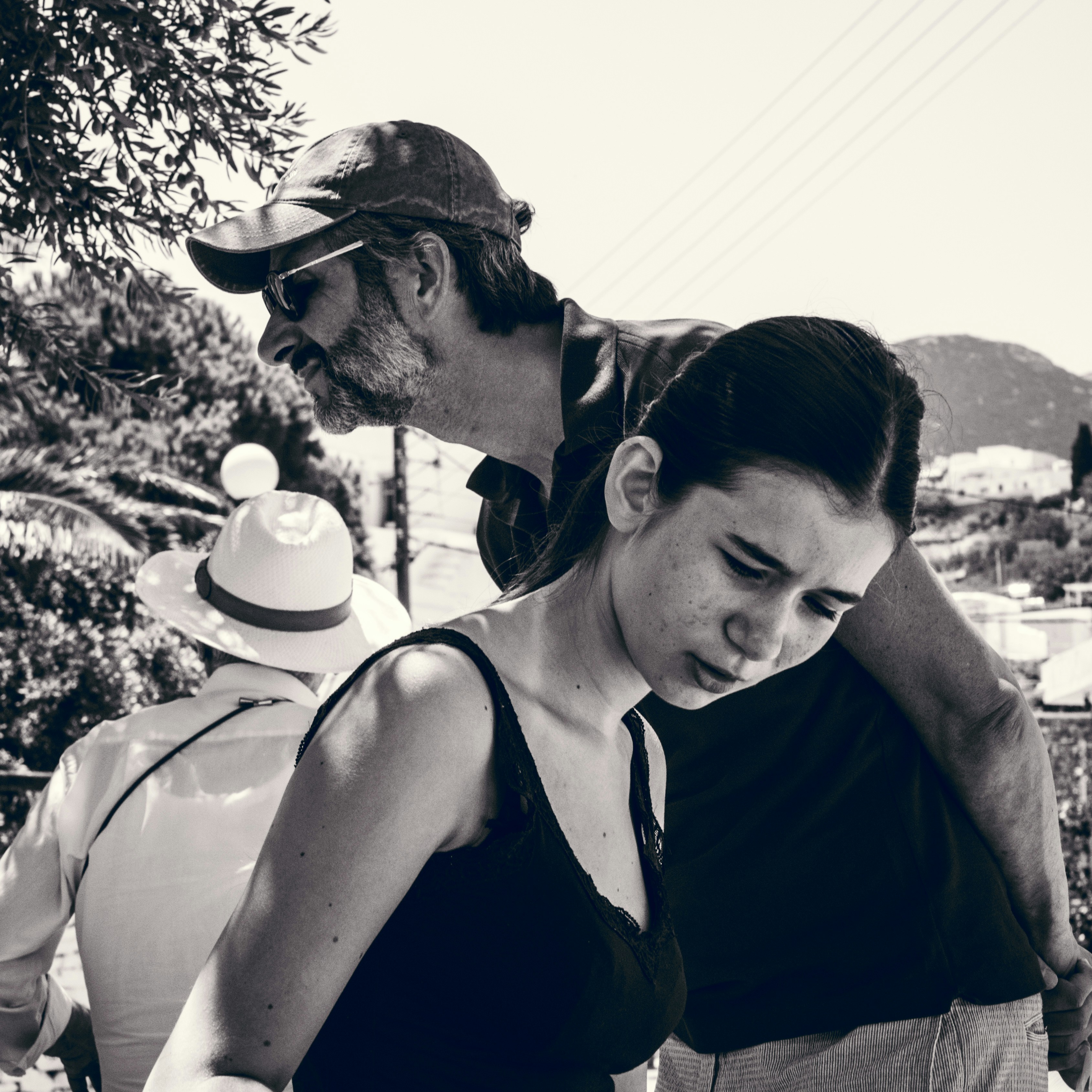 Two figures in a sunlit outdoor scene share a quiet moment, the man leaning over the woman with a protective embrace while both appear focused on something off-frame; the image emphasizes connection and subtle emotion in a candid, monochrome mood.