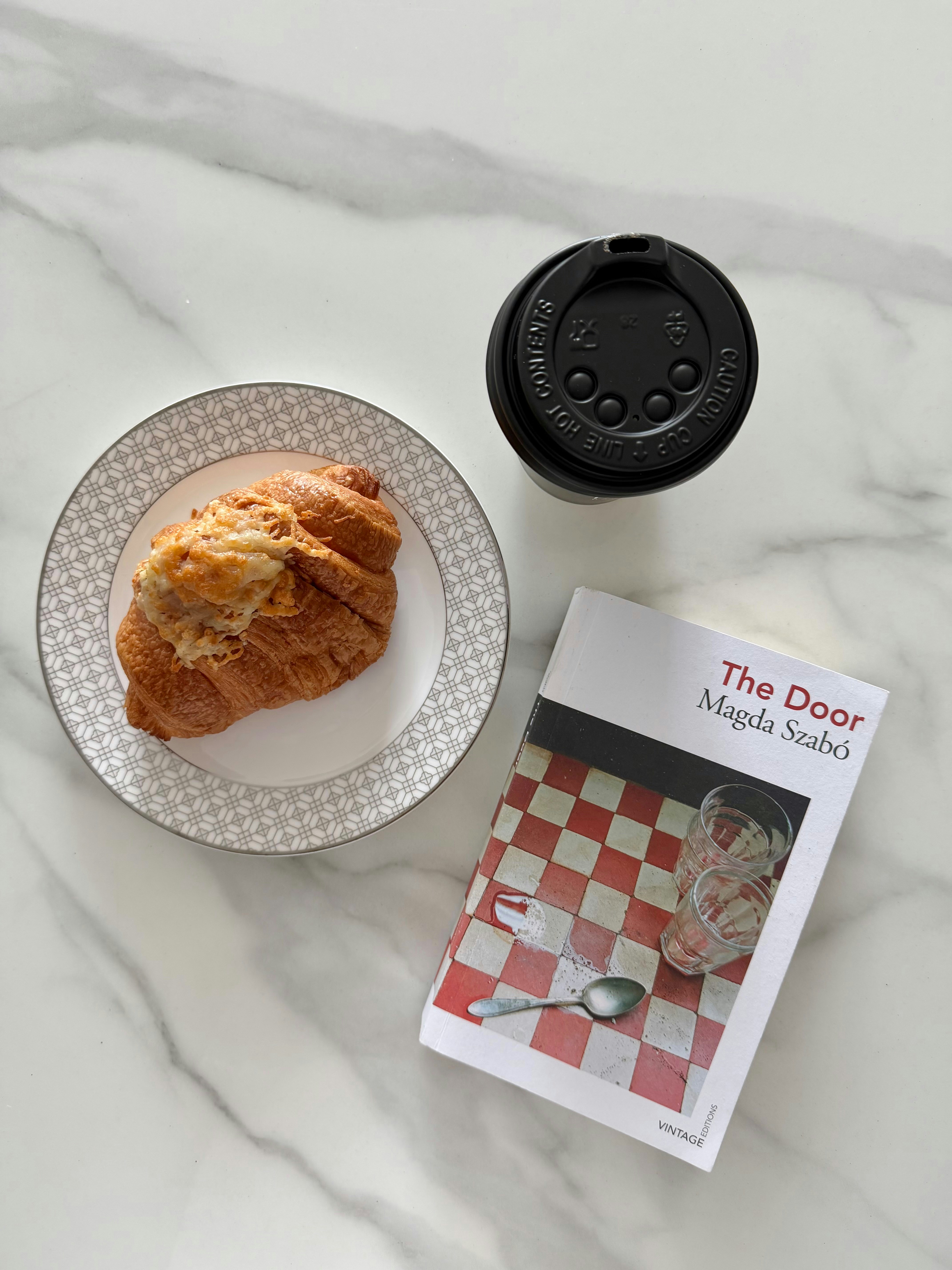 A copy of The Door by Magda Szabó beside a cup of coffee and a warm croissant on a marble table. An early morning of coffee, reading, and quiet contemplation.