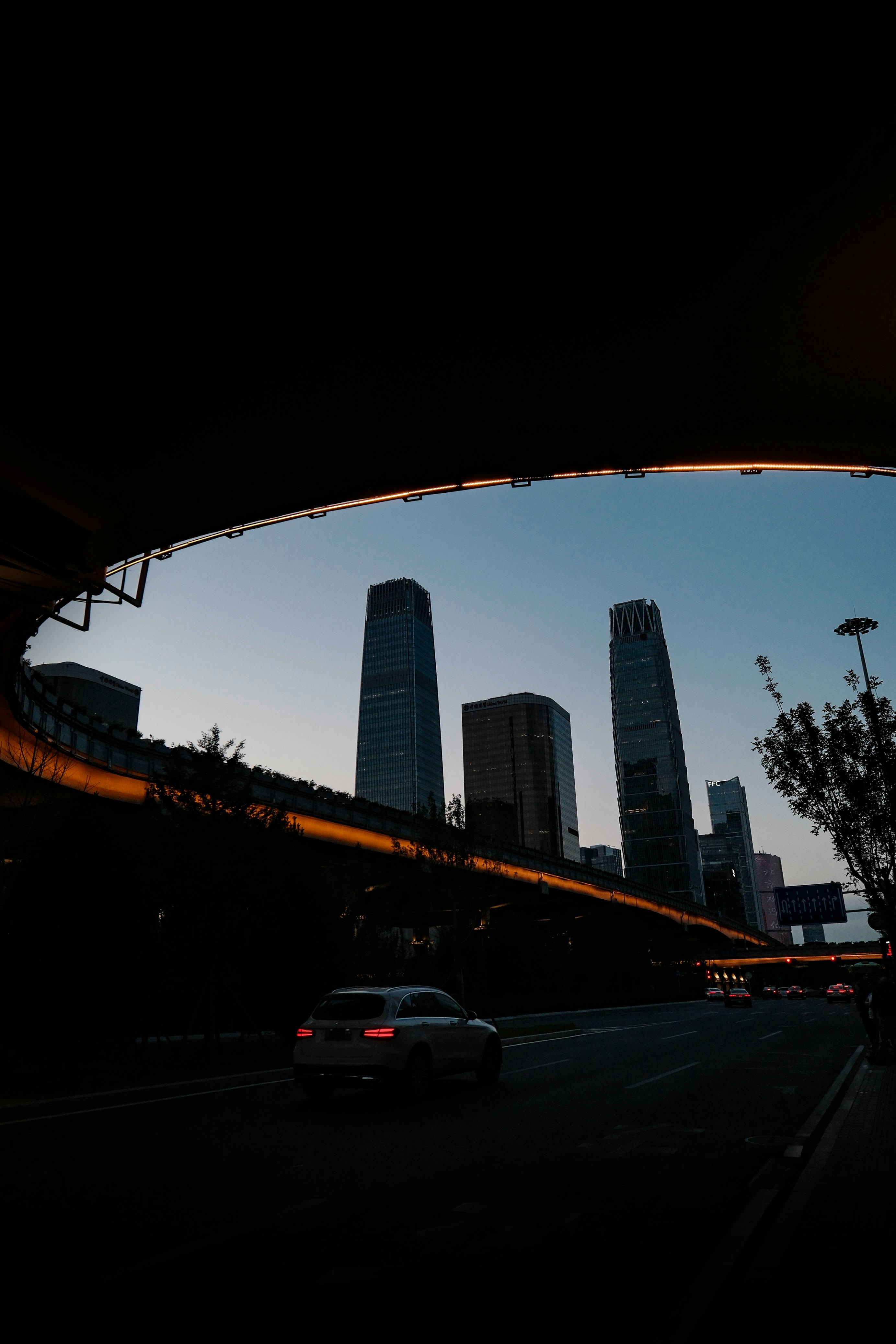 Urban highway under an illuminated overpass with tall office towers rising against dusk sky; a lone car traverses the road, creating a moody, architectural scene.