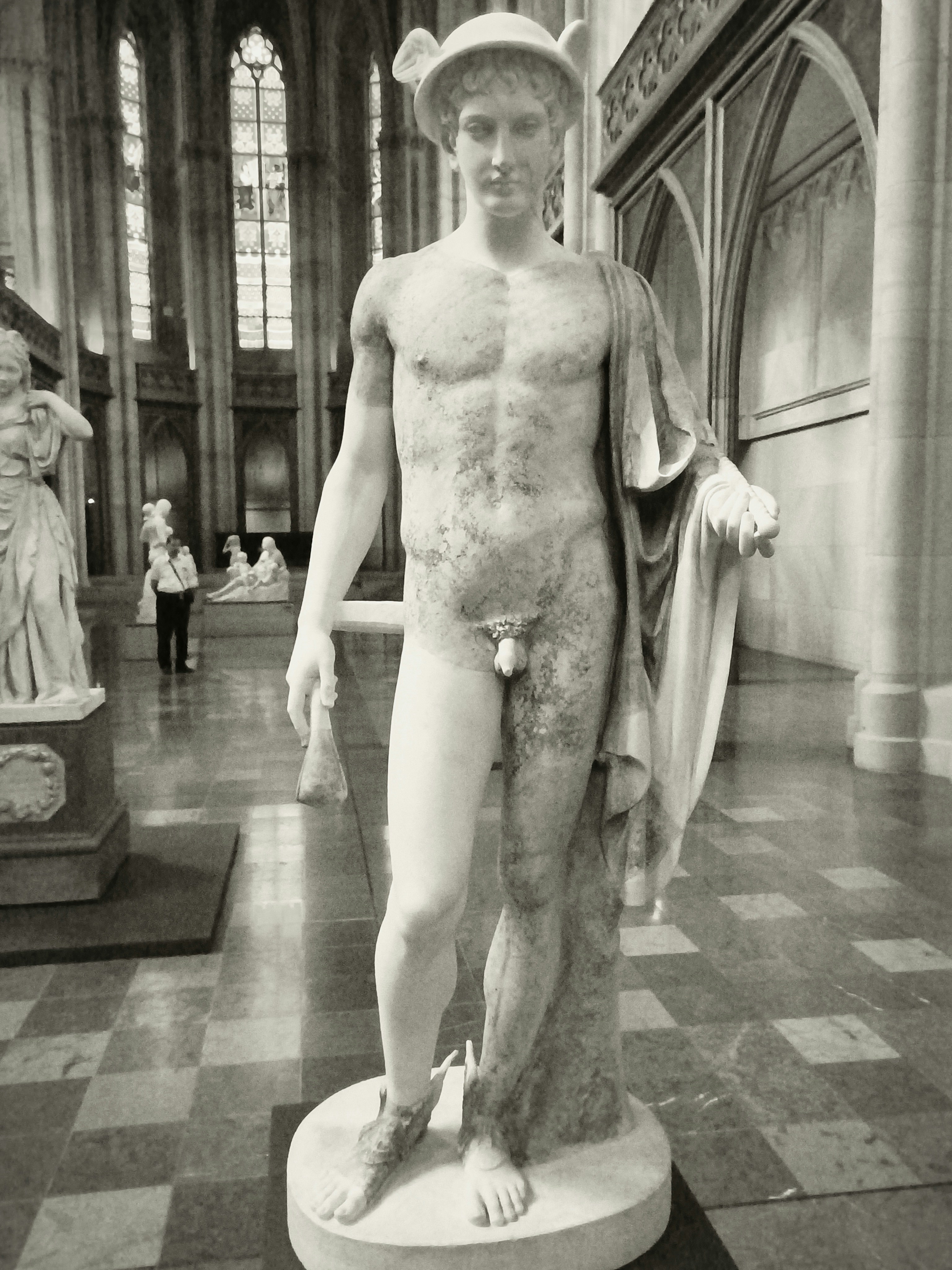 A classical statue displayed in a grand museum hall, with tall arches and other sculptures visible in the background.