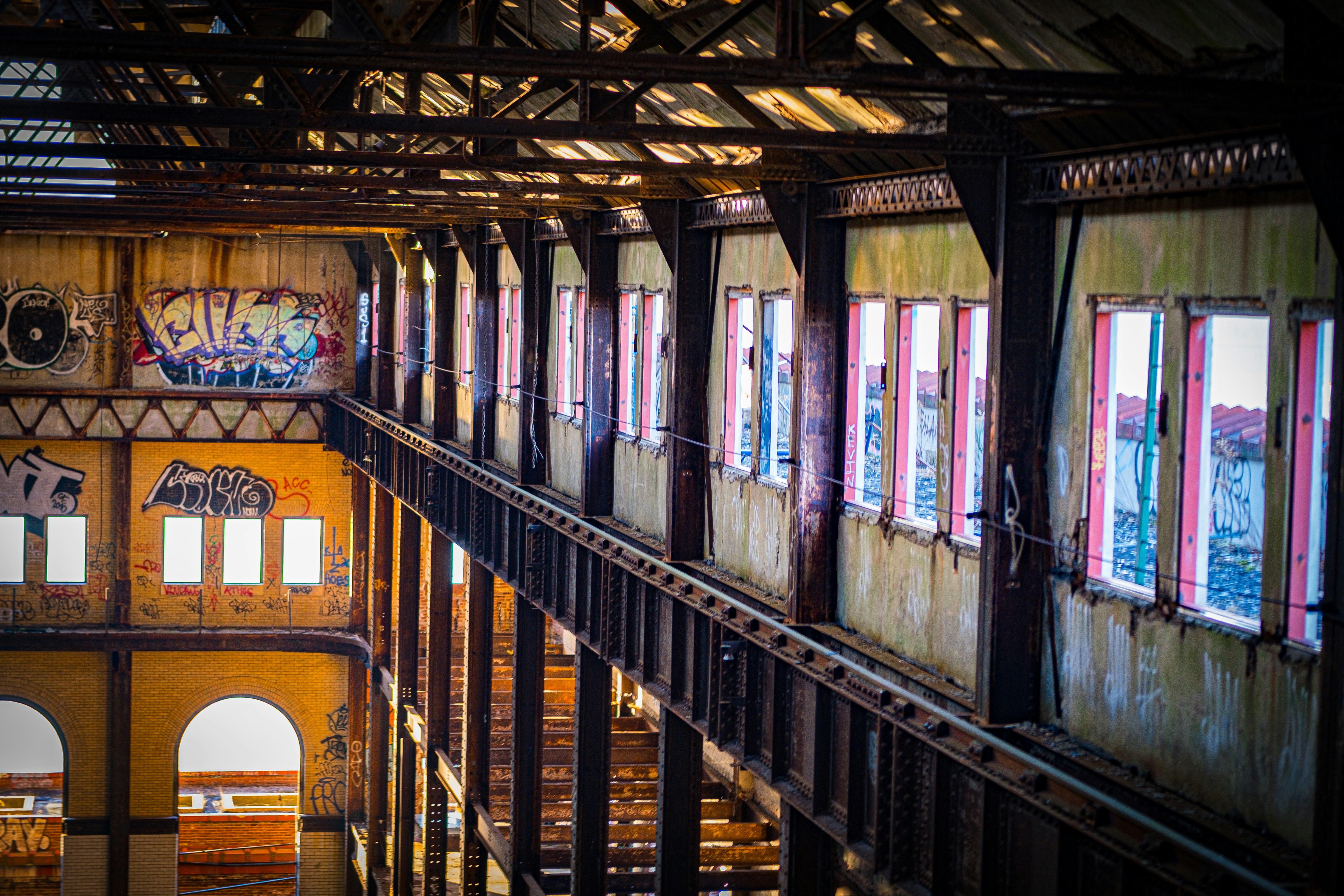 Deserted inside of a graffiti-tagged industrial building with rusted rails running along a narrow upper level and sunlit windows casting colorful light into the decay.