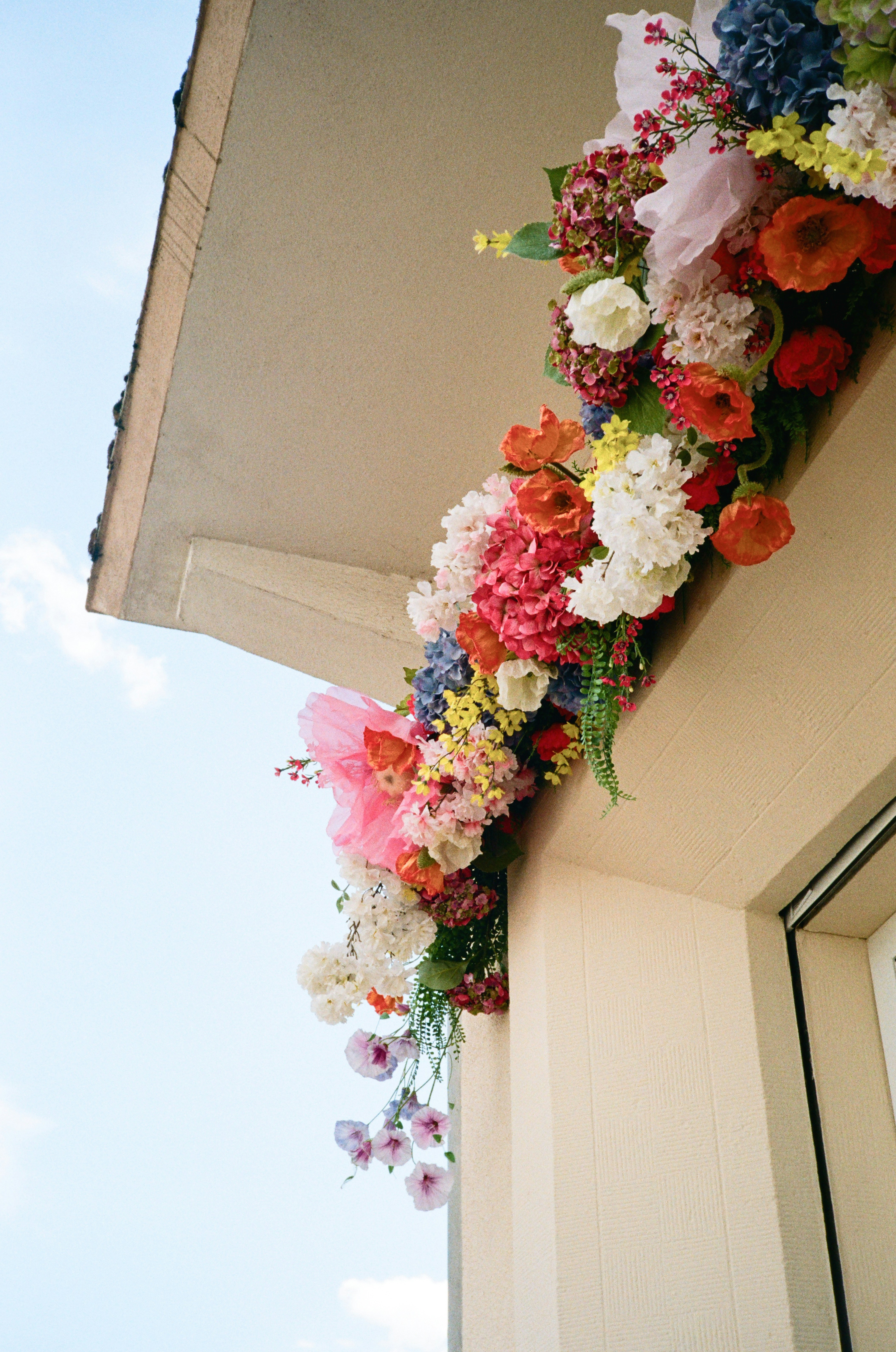 Colorful flower garland cascades along the building edge, contrasted against pale architectural surfaces and a clear blue sky.