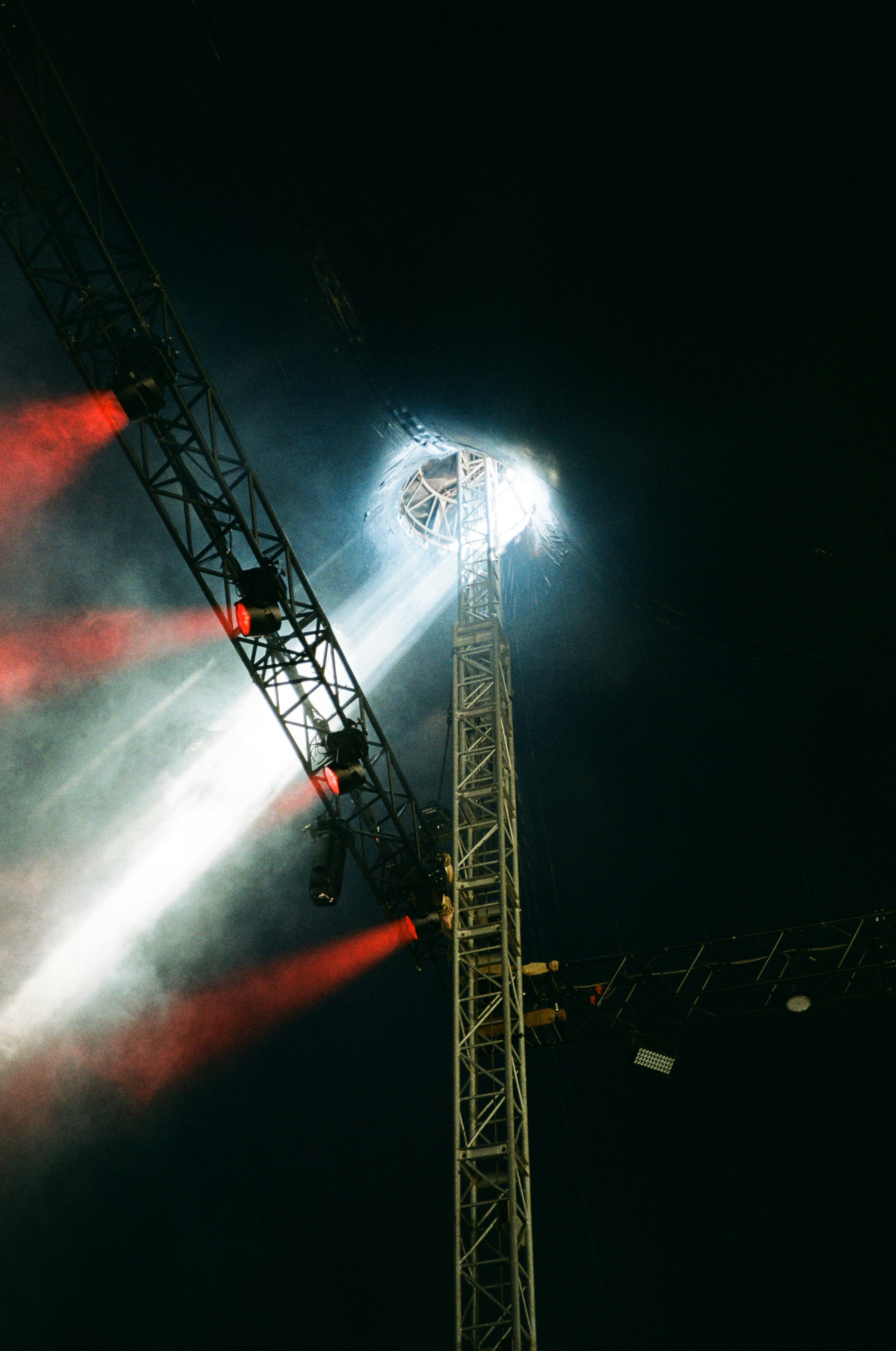 A night-time concert rig with towering crane structure and dramatic spotlights carving white and red beams across the frame. The metallic tower anchors the composition, while smoky atmosphere adds depth and motion.
