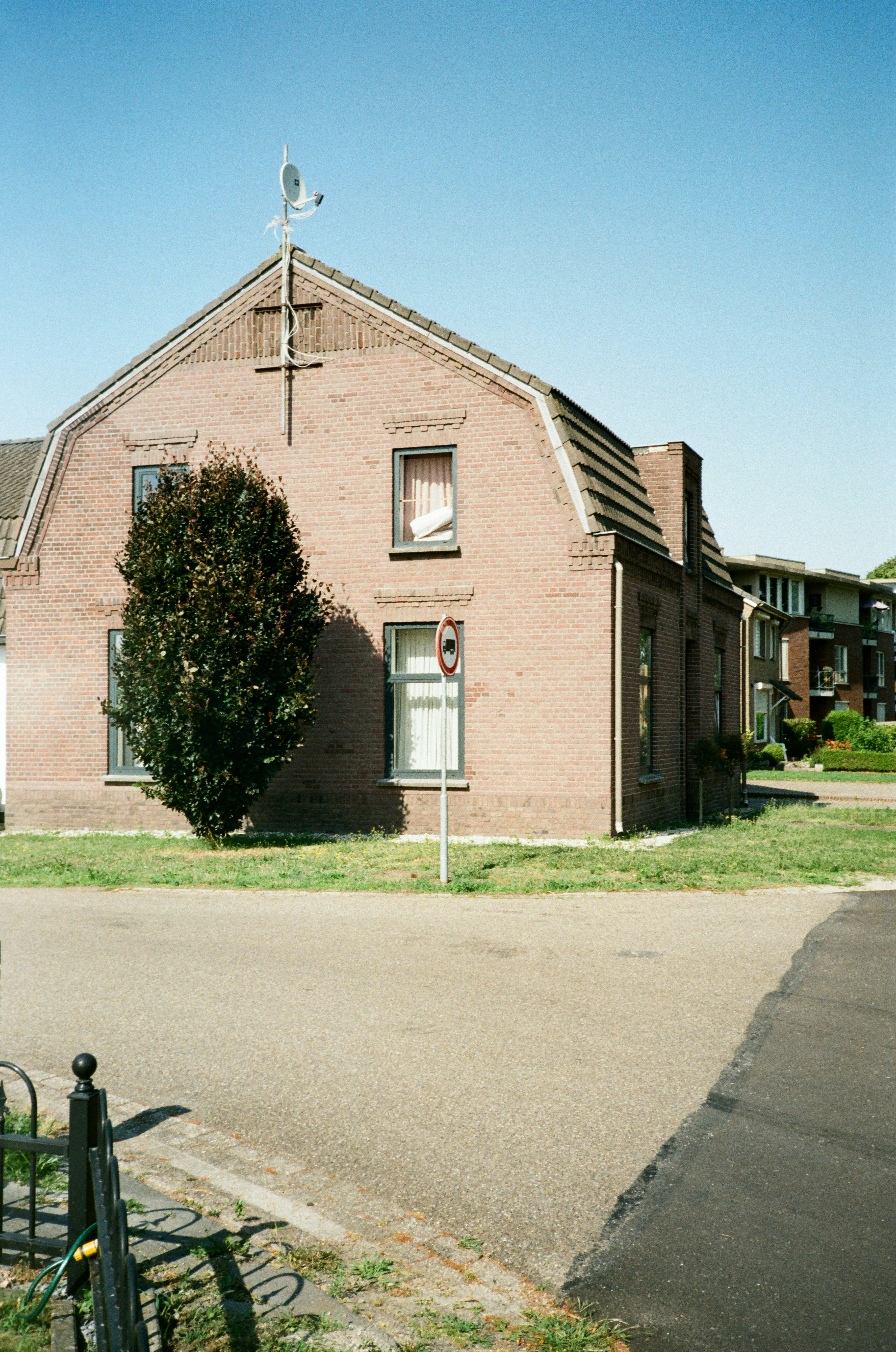 Brick house with a tree in front and a street sign, set in a quiet suburban street; warm daylight highlights the building and surrounding pavement.