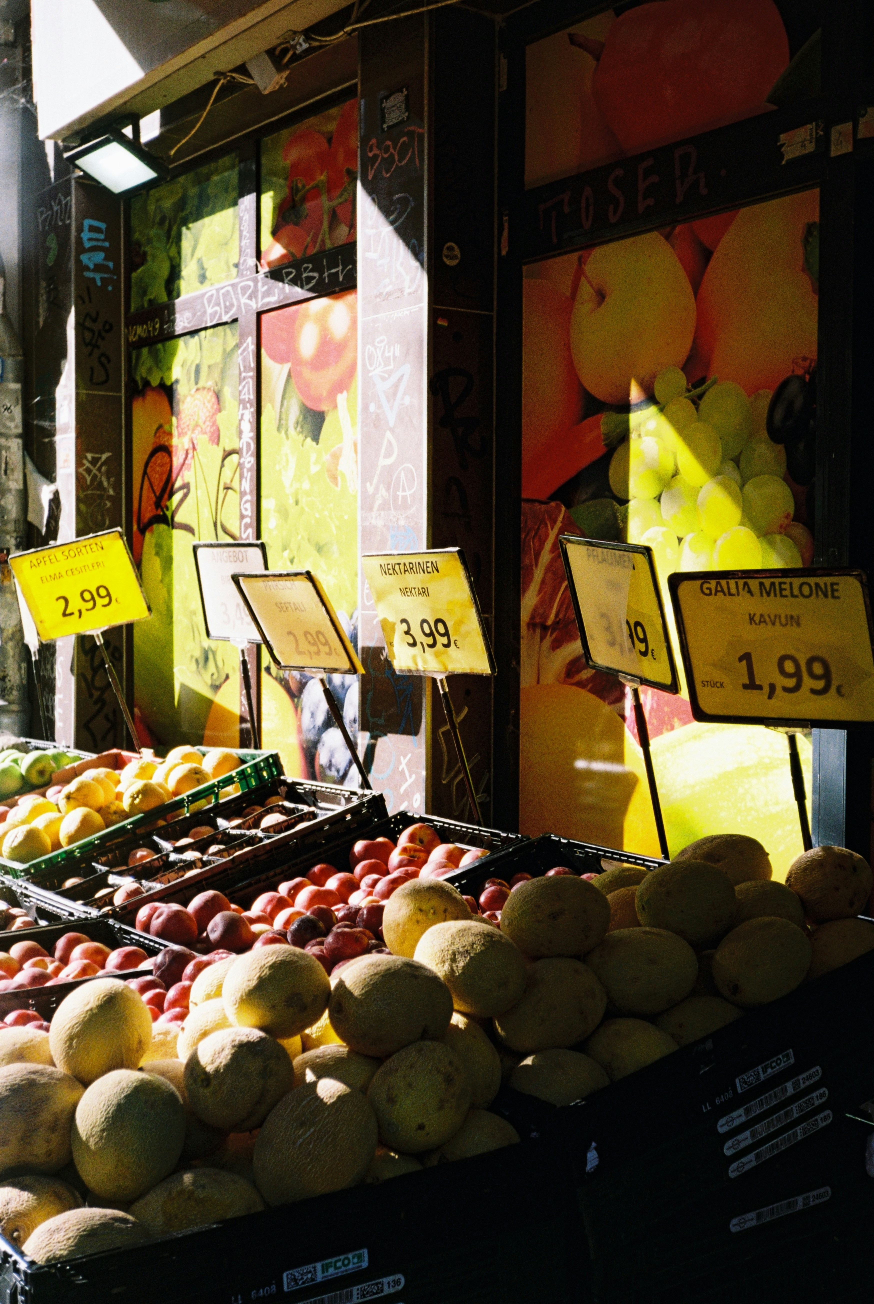 Colorful fruit stalls glow under graffiti-adorned shopfronts with illuminated price signs in a sunlit market alley.