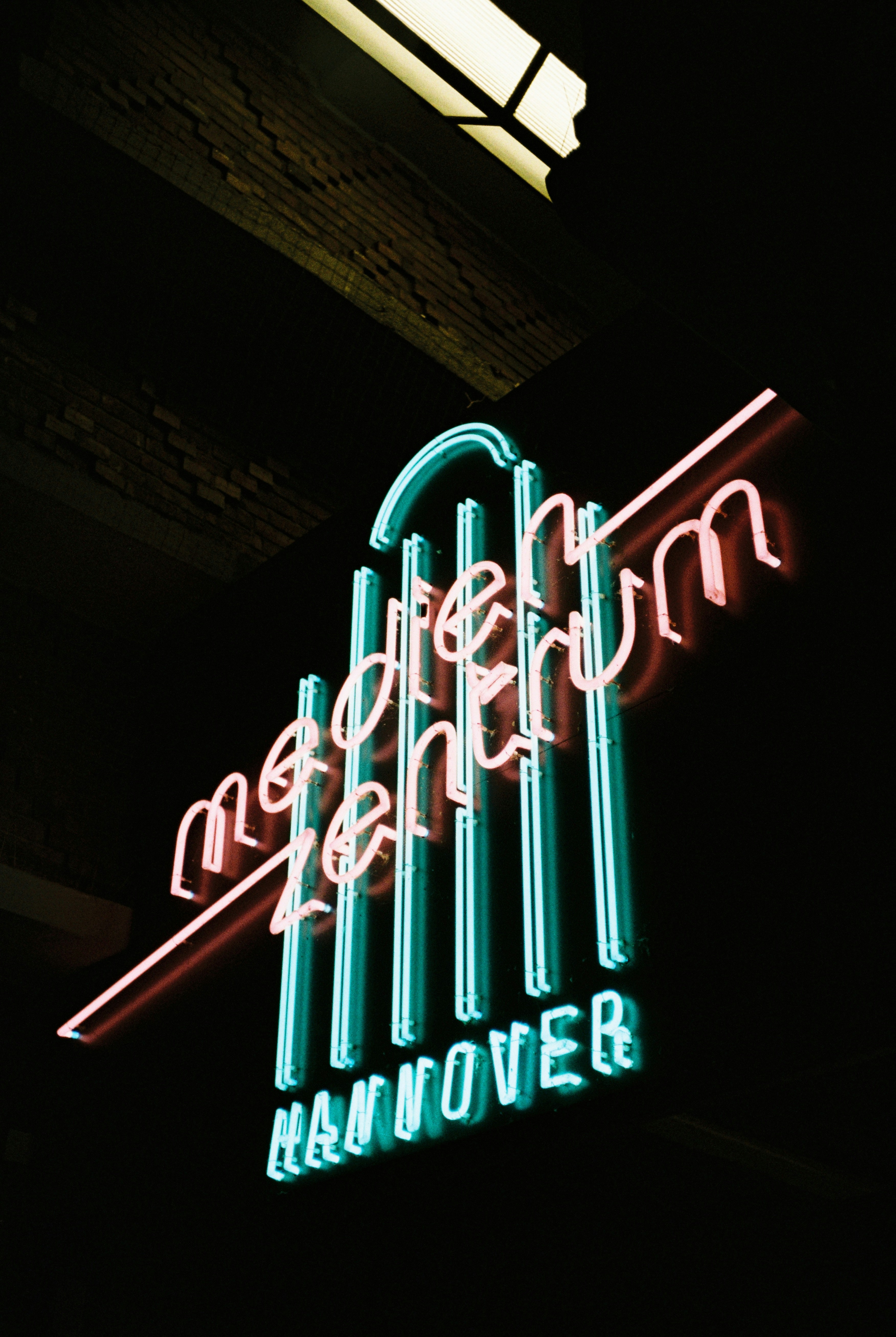 Neon sign in teal and pink glows against a dark urban backdrop, the wordmark slightly tilted as vertical tubes form a stylized letter pattern. A moody, late-night city scene with emphasis on luminous signage.