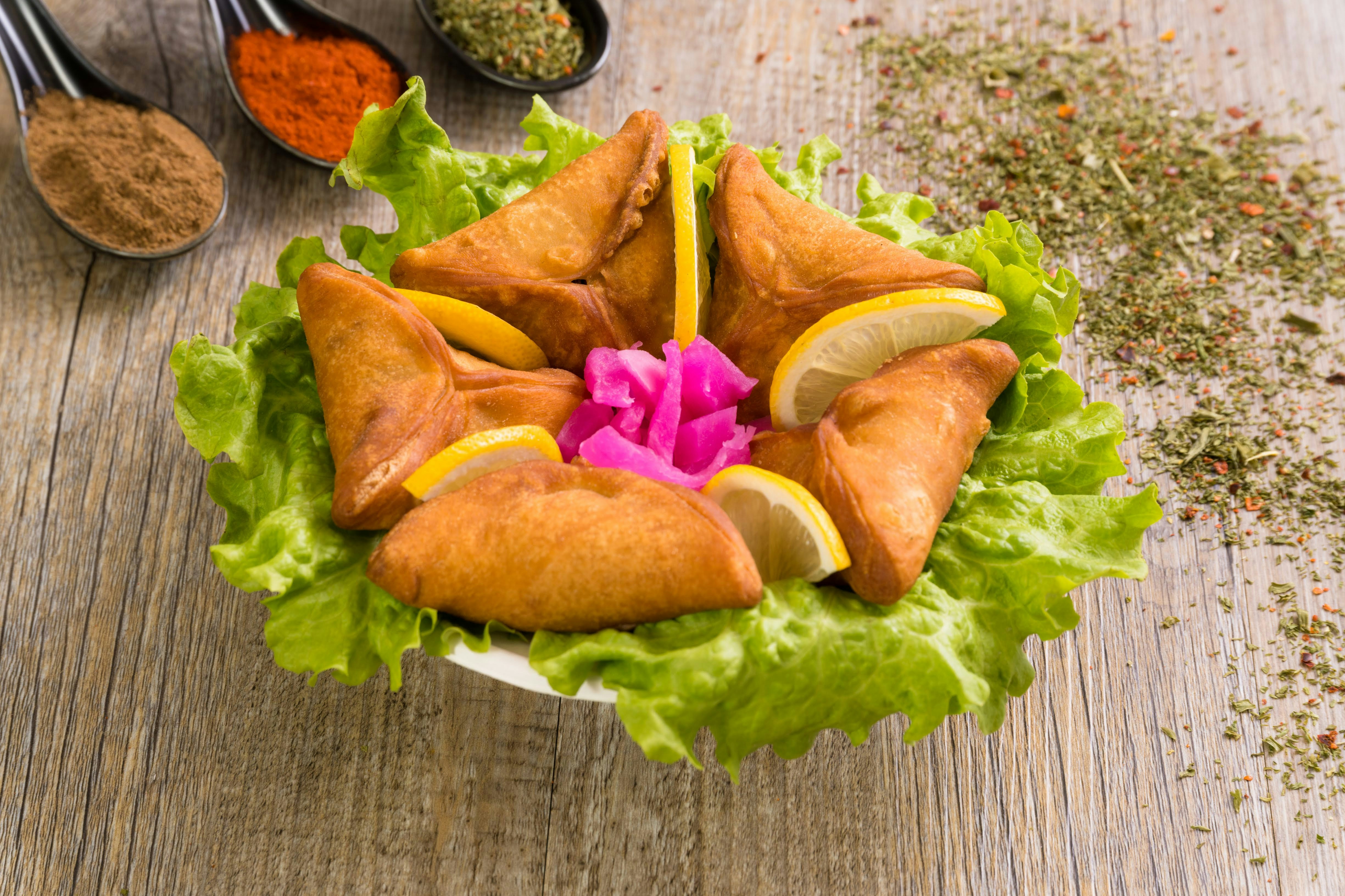 Plate of golden samosas arranged on lettuce with lemon wedges, set on a rustic wooden table; a garnish of colorful spices visible in the background. This is a food presentation shot.
