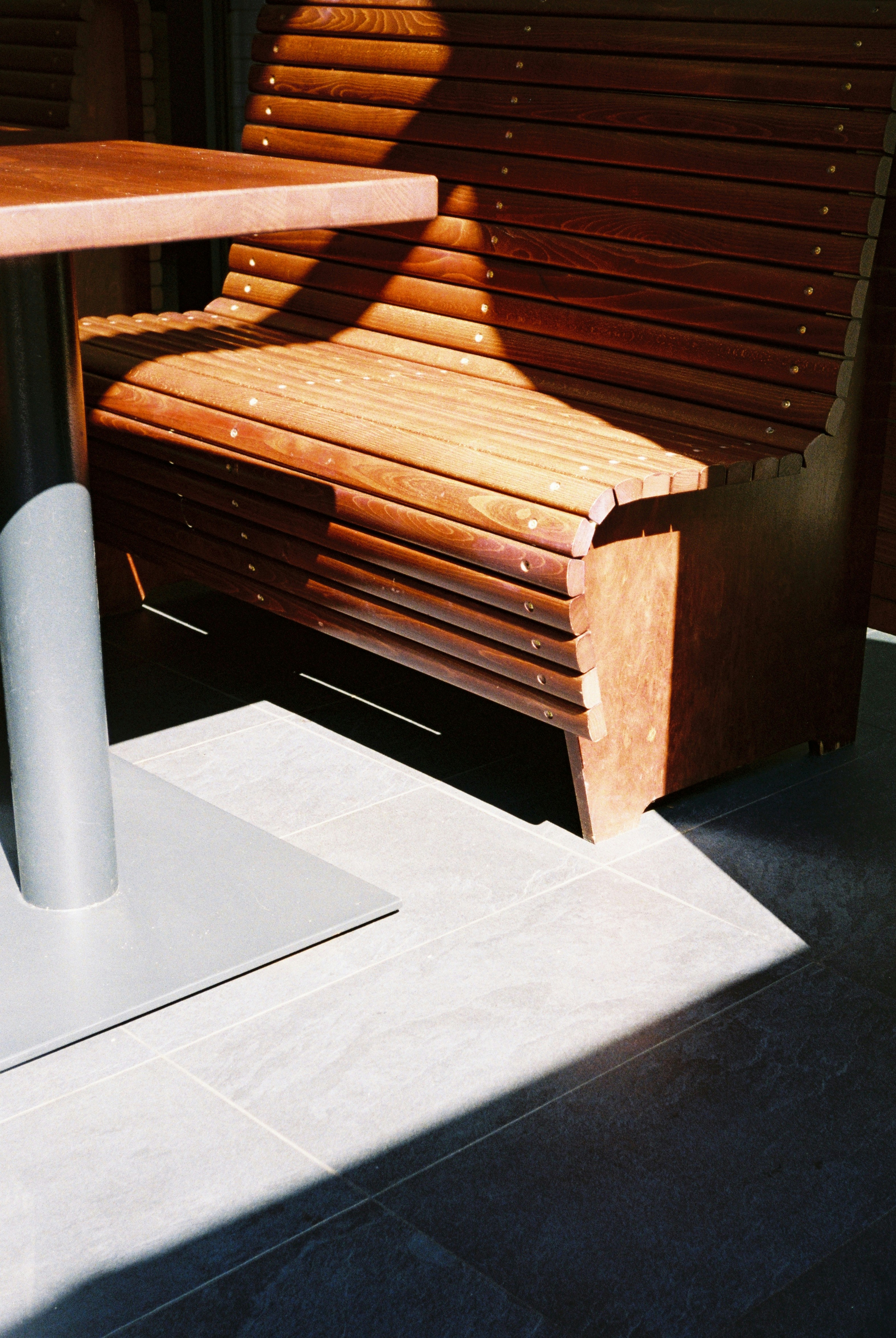 Sunlit wooden slatted bench under a table in a shaded patio; warm tones and strong shadows suggest midday sun.