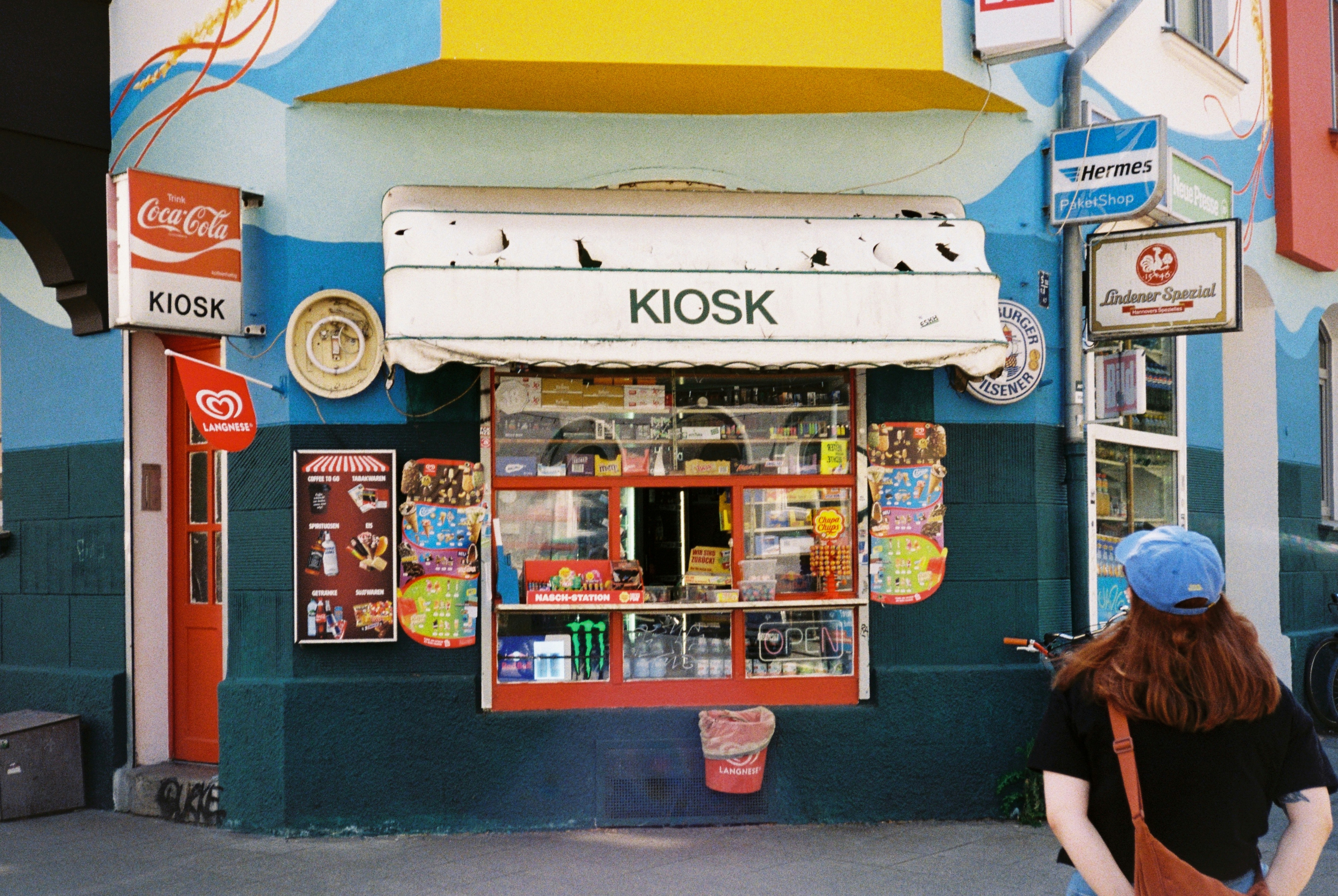 Vibrant kiosk storefront with a white awning labeled KIOSK, flanked by colorful signage and a passerby in the foreground.