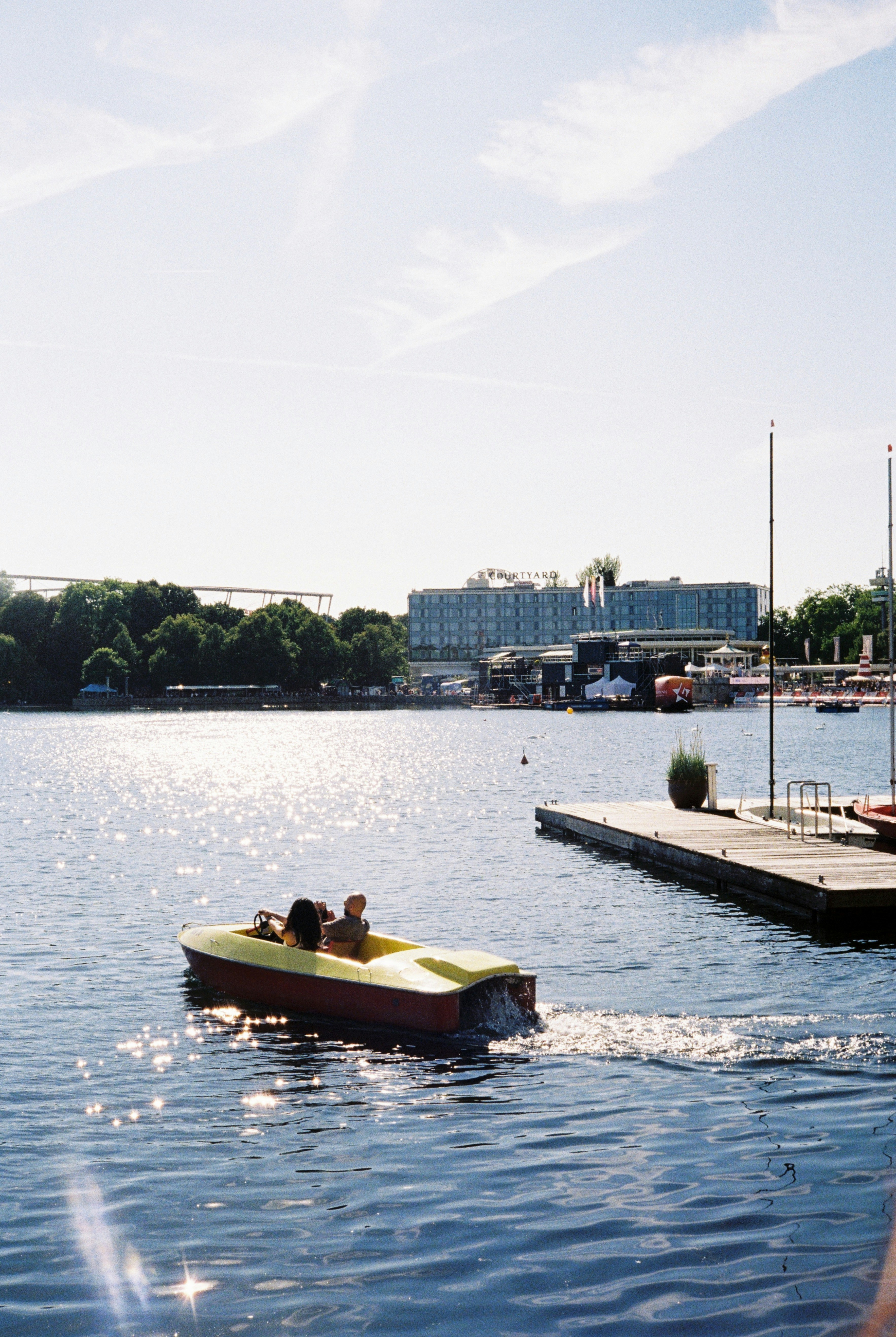 Sunlit lake scene with a small yellow boat carrying two passengers cruising past a marina, reflections glimmering on the water and a distant hotel façade overlooking the shore.