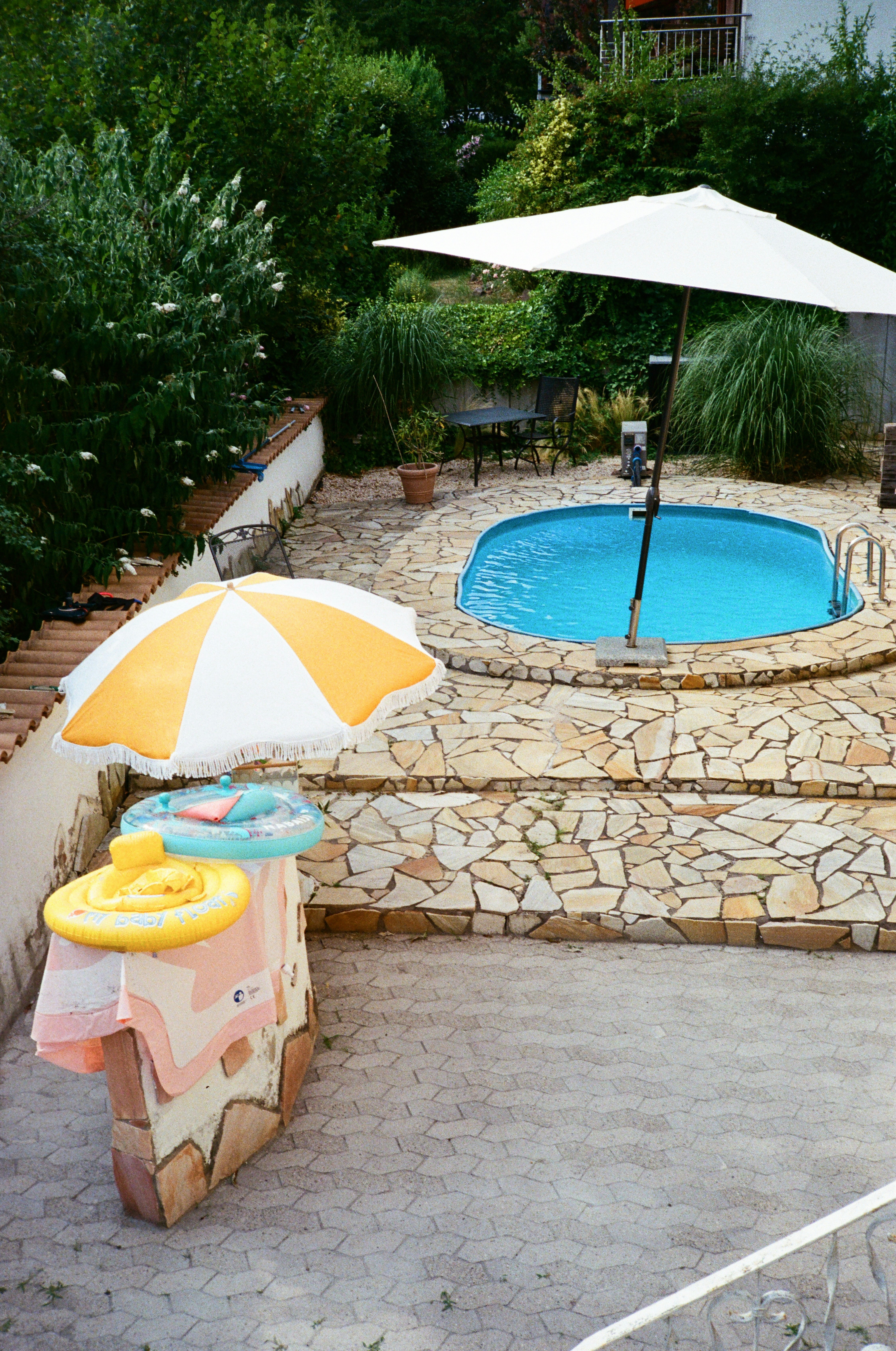A sunlit backyard features a curved blue pool surrounded by a stone patio and lush greenery, with a white umbrella casting shade over the seating area and a yellow-striped umbrella near a decorative plaza.