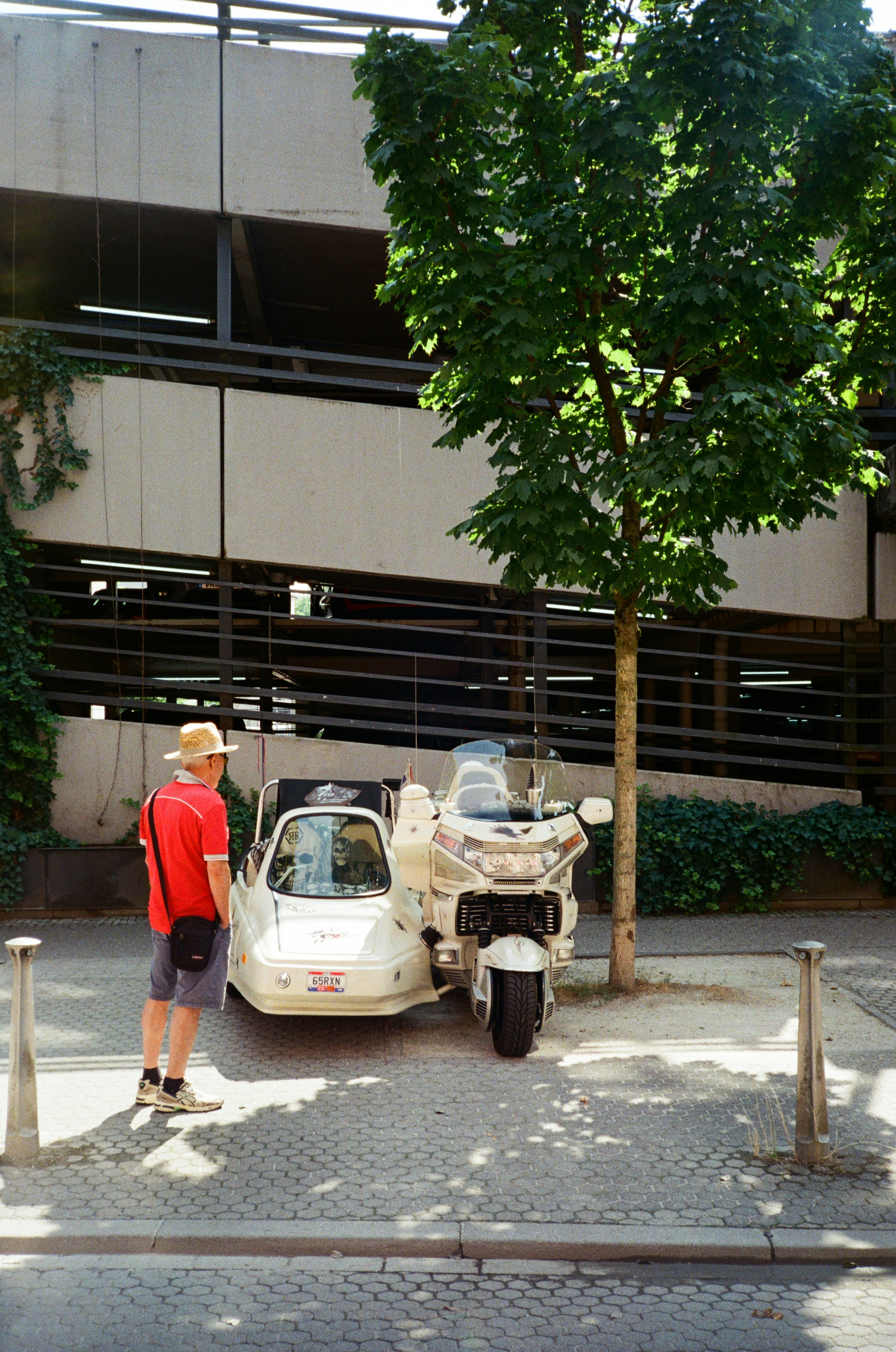 A man in a red shirt with a hat stands beside a white motorcycle and sidecar in a cobblestone lot, with a multi-level parking structure and trees in the background. The scene is outdoors and well-lit.