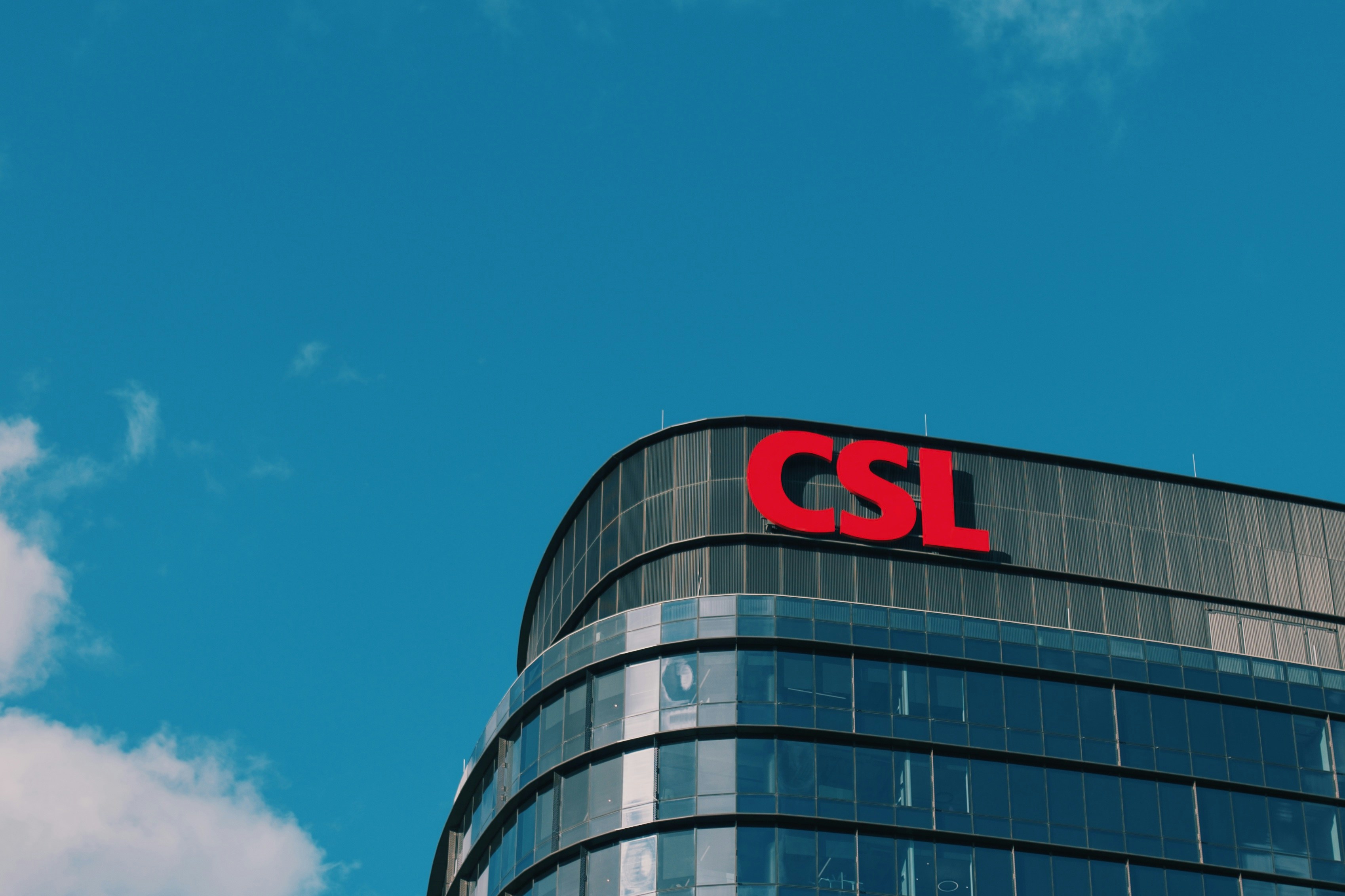 Crested CSL logo atop a curved glass façade against a bright blue sky; architectural detail highlighting modern office building.