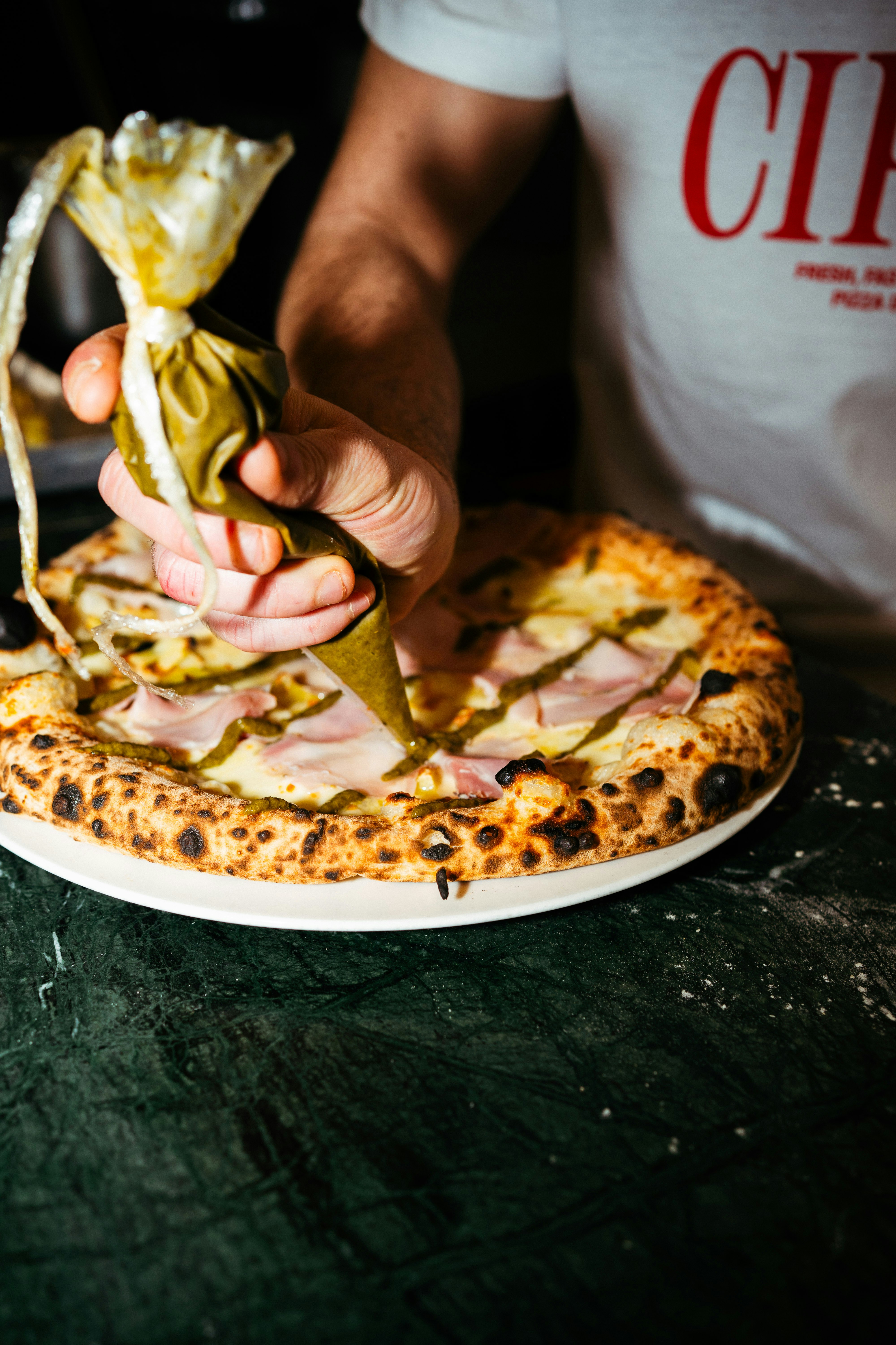 Close-up of a person squeezing sauce onto a hot wood-fired pizza, the crust puffy with char and melted cheese visible. A hands-on cooking moment captured with focus on texture and action.