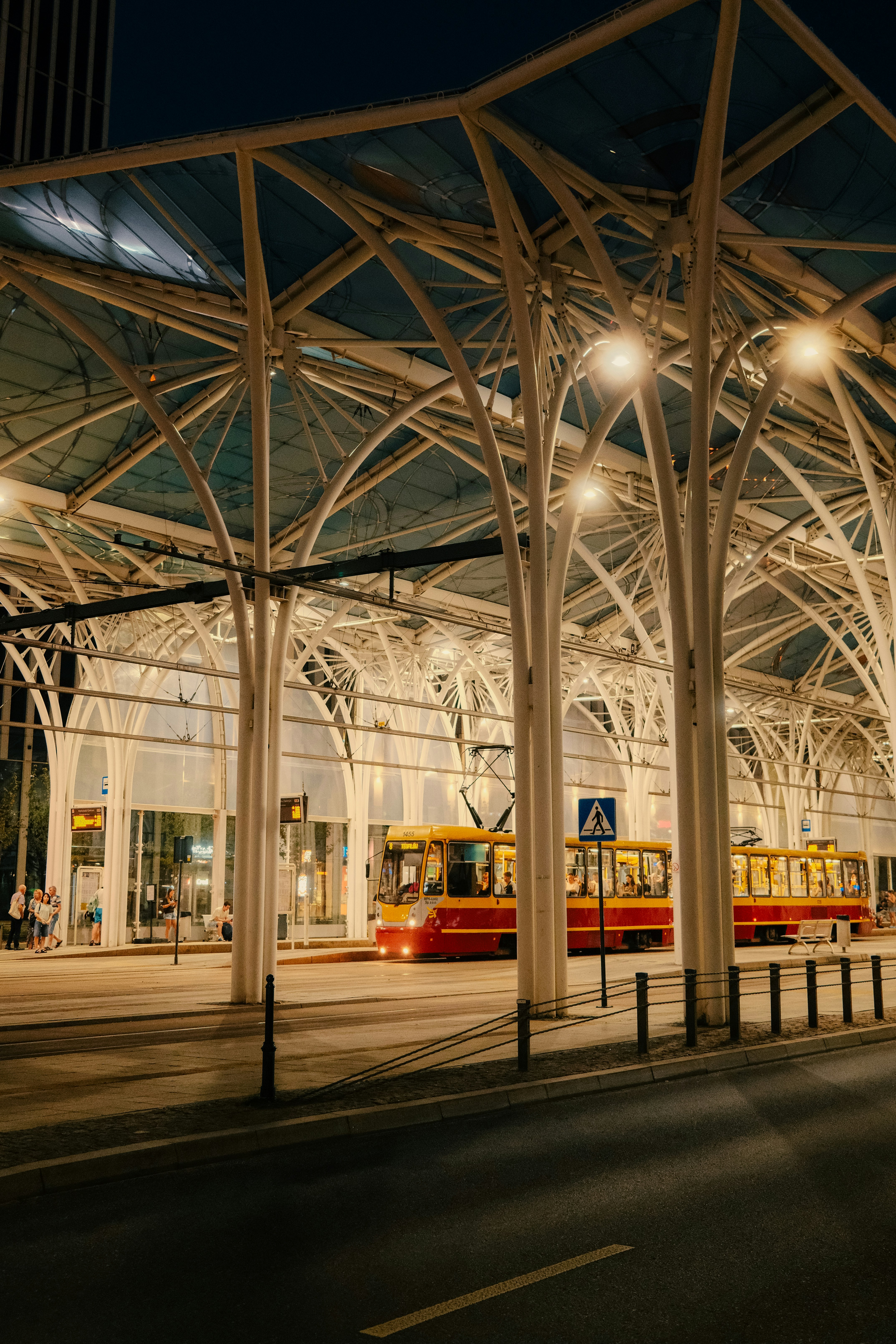 Night scene of a red and yellow tram stopping at a modern glass-and-steel transit canopy, with bright internal lights casting warm tones on the platform and nearby pedestrians.