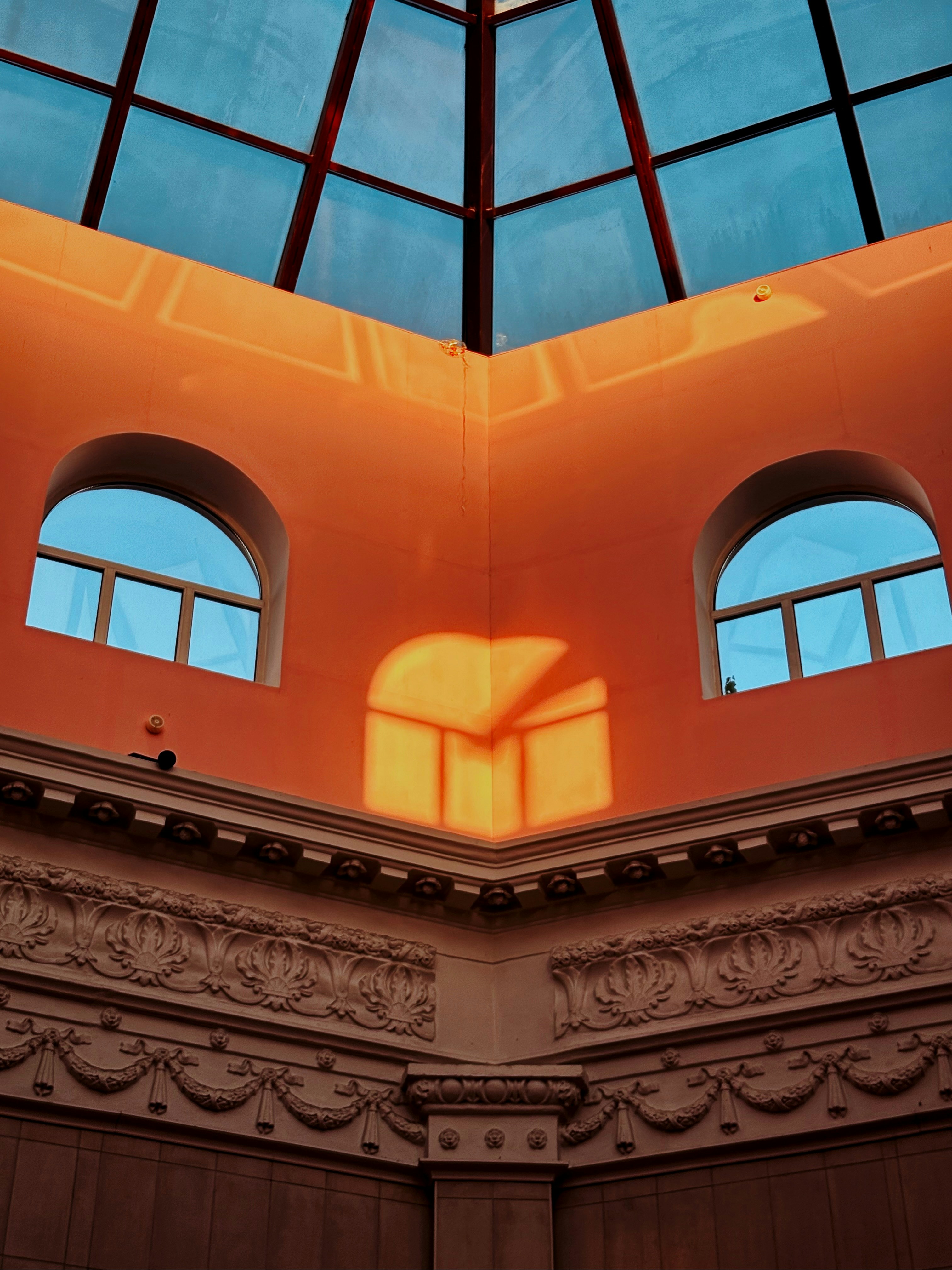 Warm orange walls rise to a glass ceiling, with arched windows casting cool blue light; a soft shadowed balcony moldings frame the lower edge of the scene.