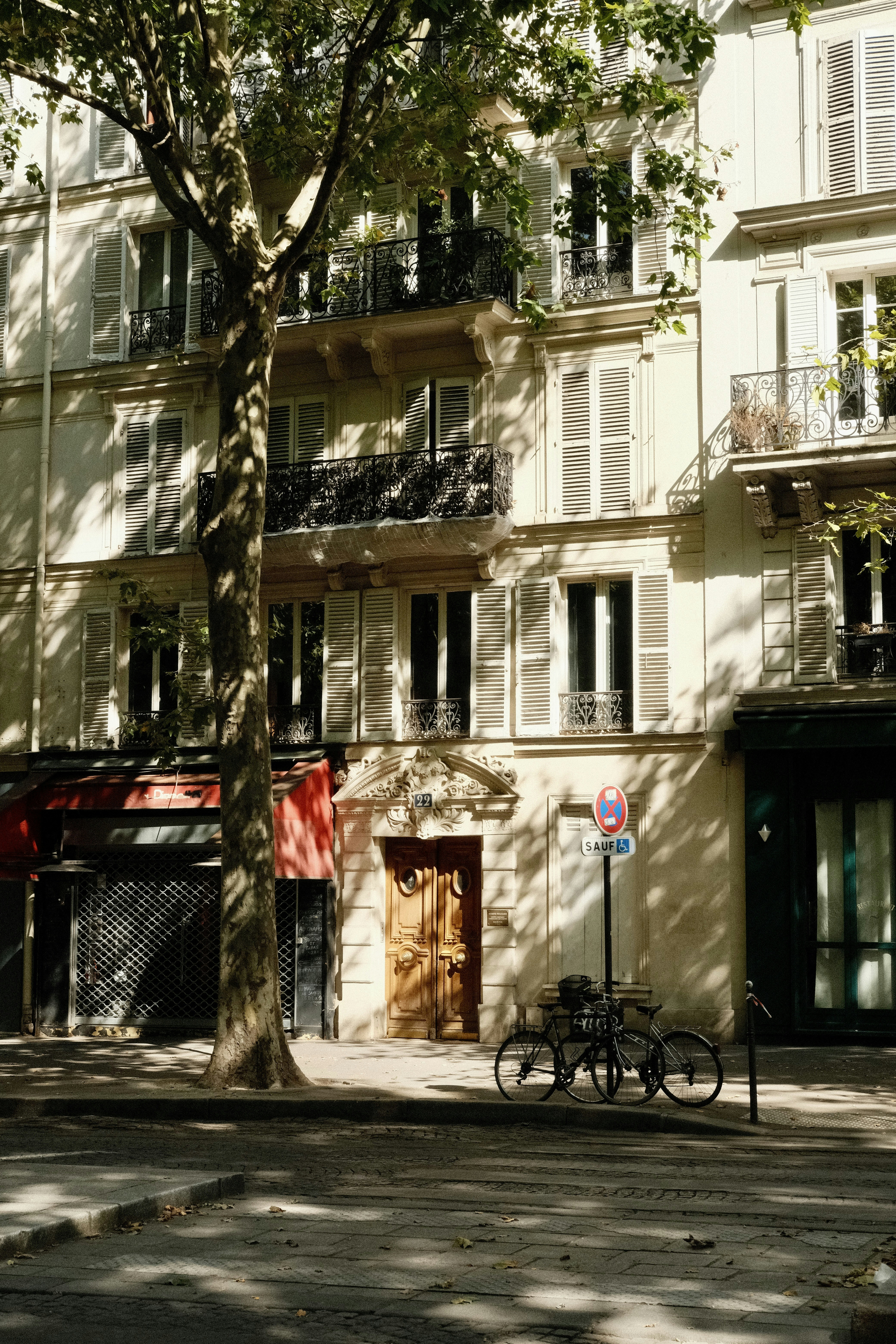 Sun-dappled street scene featuring a classic Parisian building with shuttered windows, a wooden door, a bicycle, and a tree casting patterned shadows on the façade.