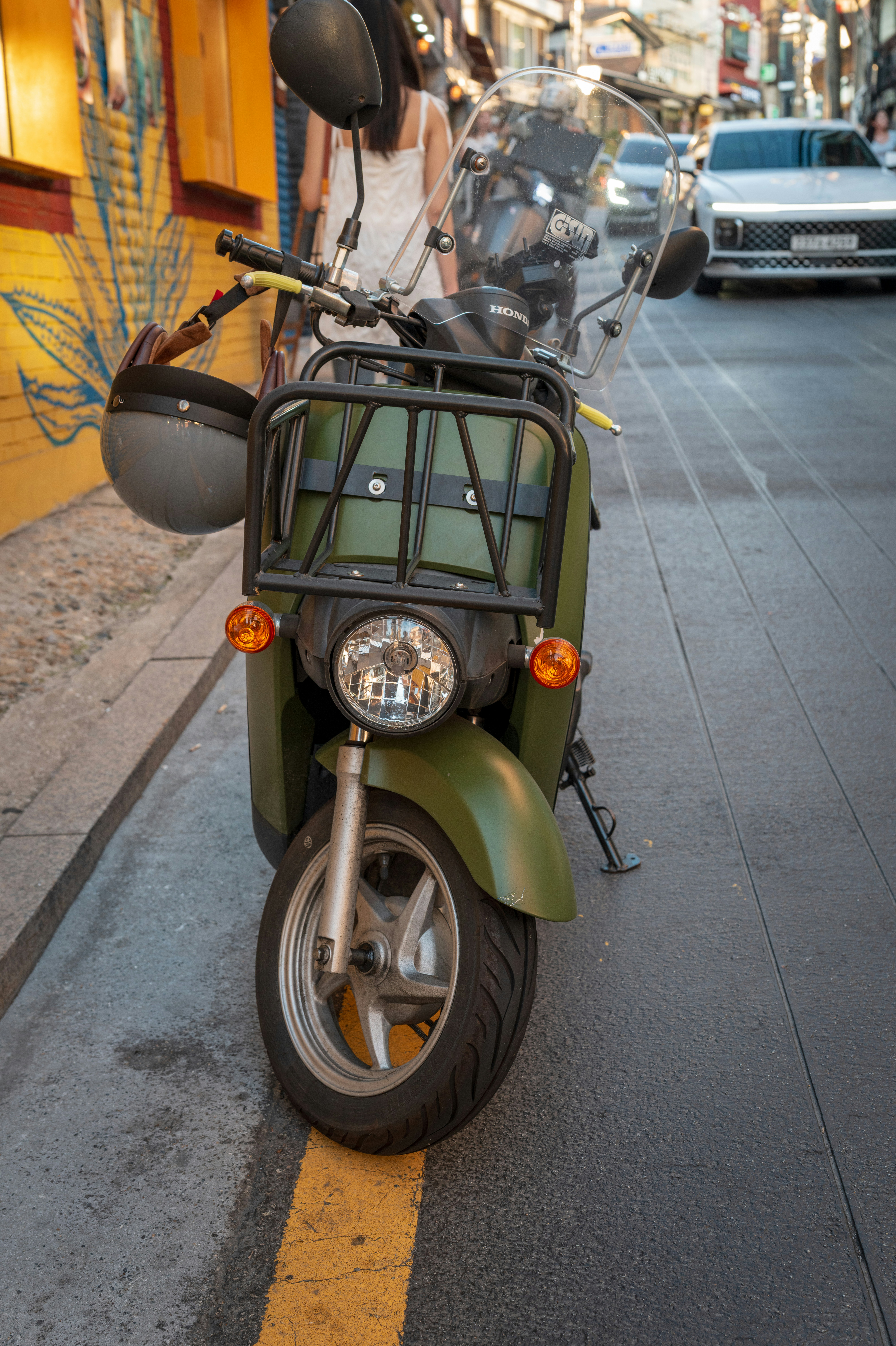 Green scooter with a protective front rack sits on a curb-side street, a helmet attached to the side, urban storefronts and a pedestrian in the background. A city portrait of a compact, utilitarian ride.
