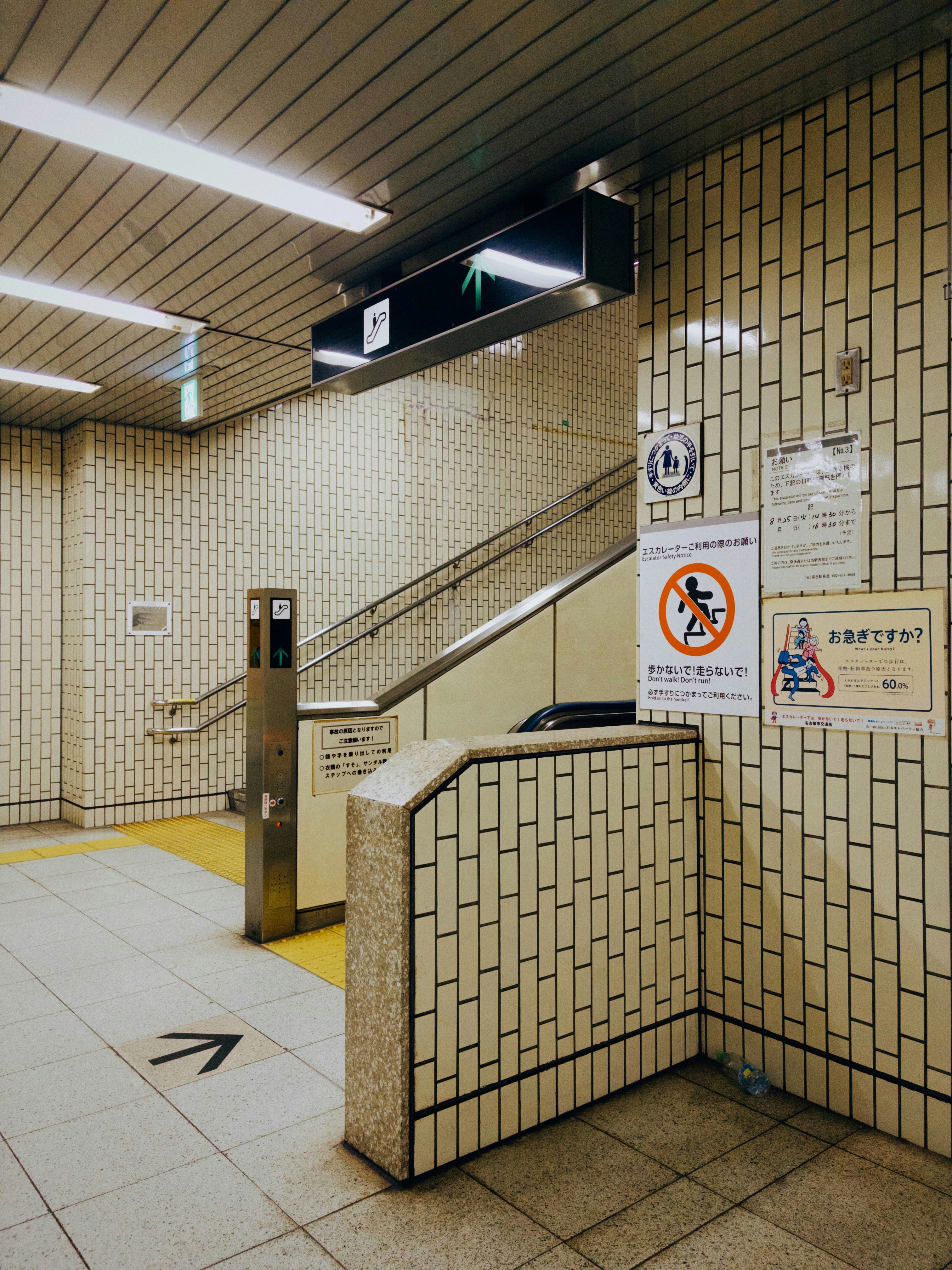 Underscored subway passage with tiled walls, a turnstile, and a staircase in the background; signs and a yellow-tiled floor create a calm, utilitarian scene.