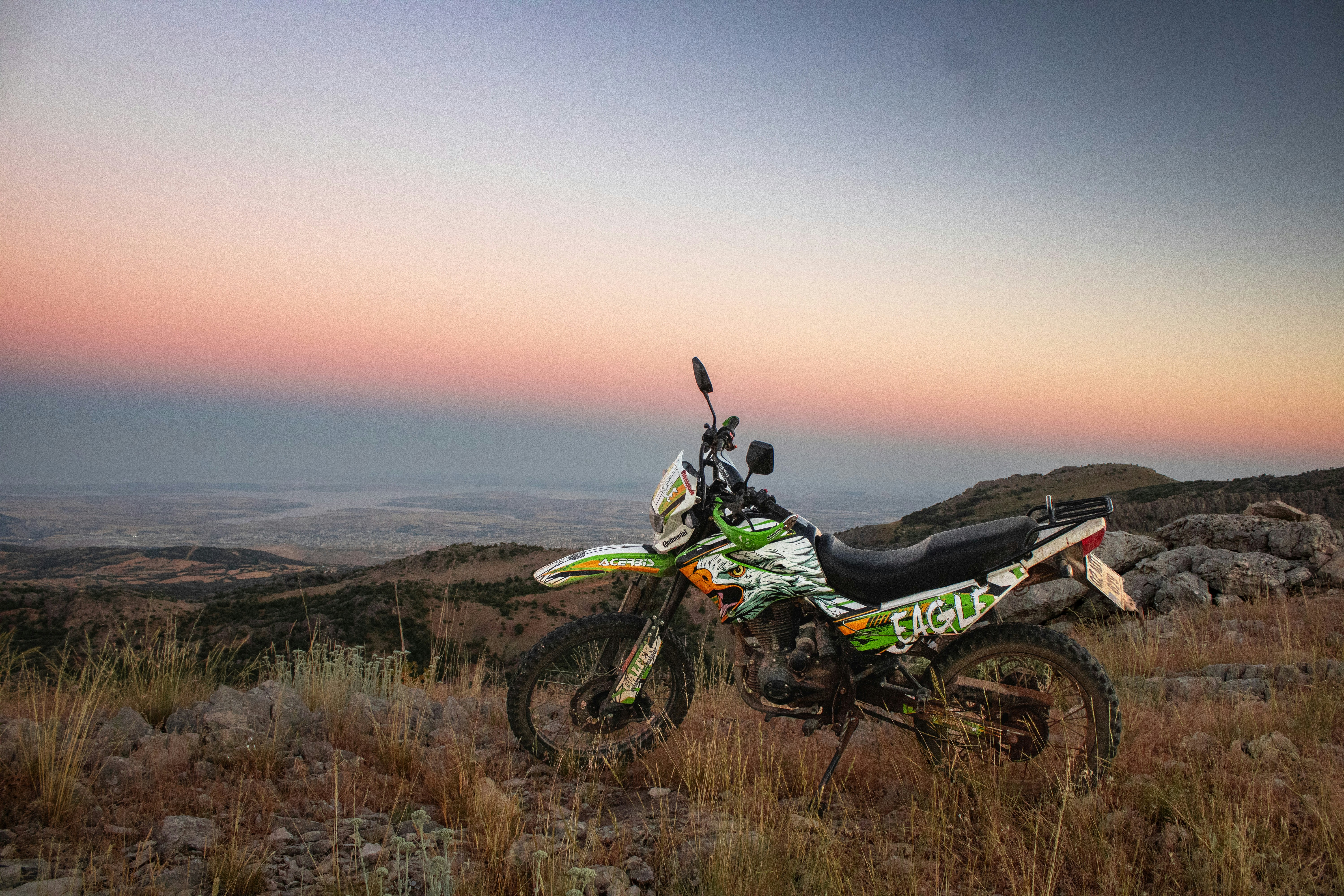 Off-road motorcycle parked on a rocky hillside at sunset with a gradual desert valley beyond.