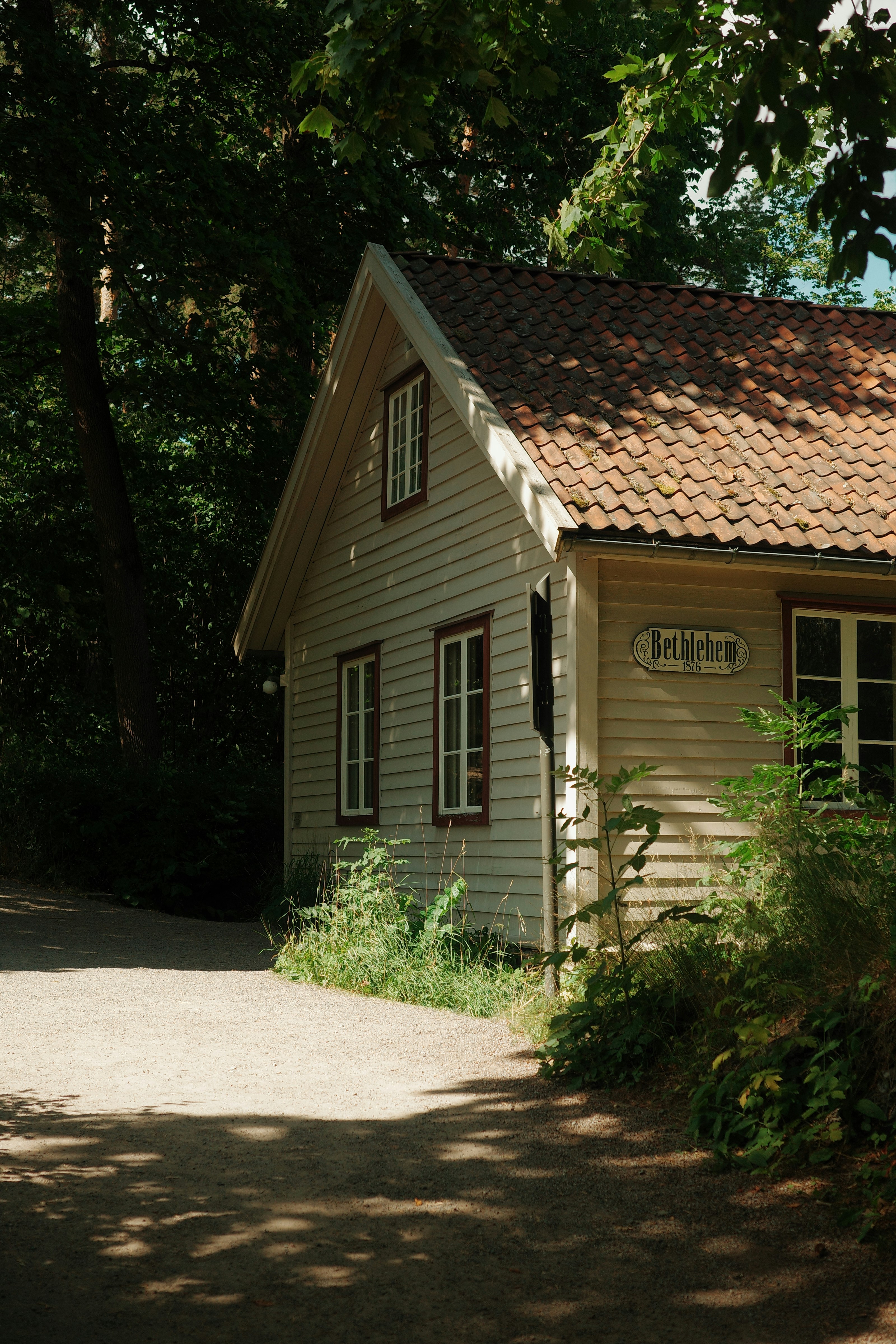 Sunlit wooden cottage with a tile roof sits beside a dirt path; overhanging trees cast dappled shadows on the side, and a small sign decorates the wall near the windows.